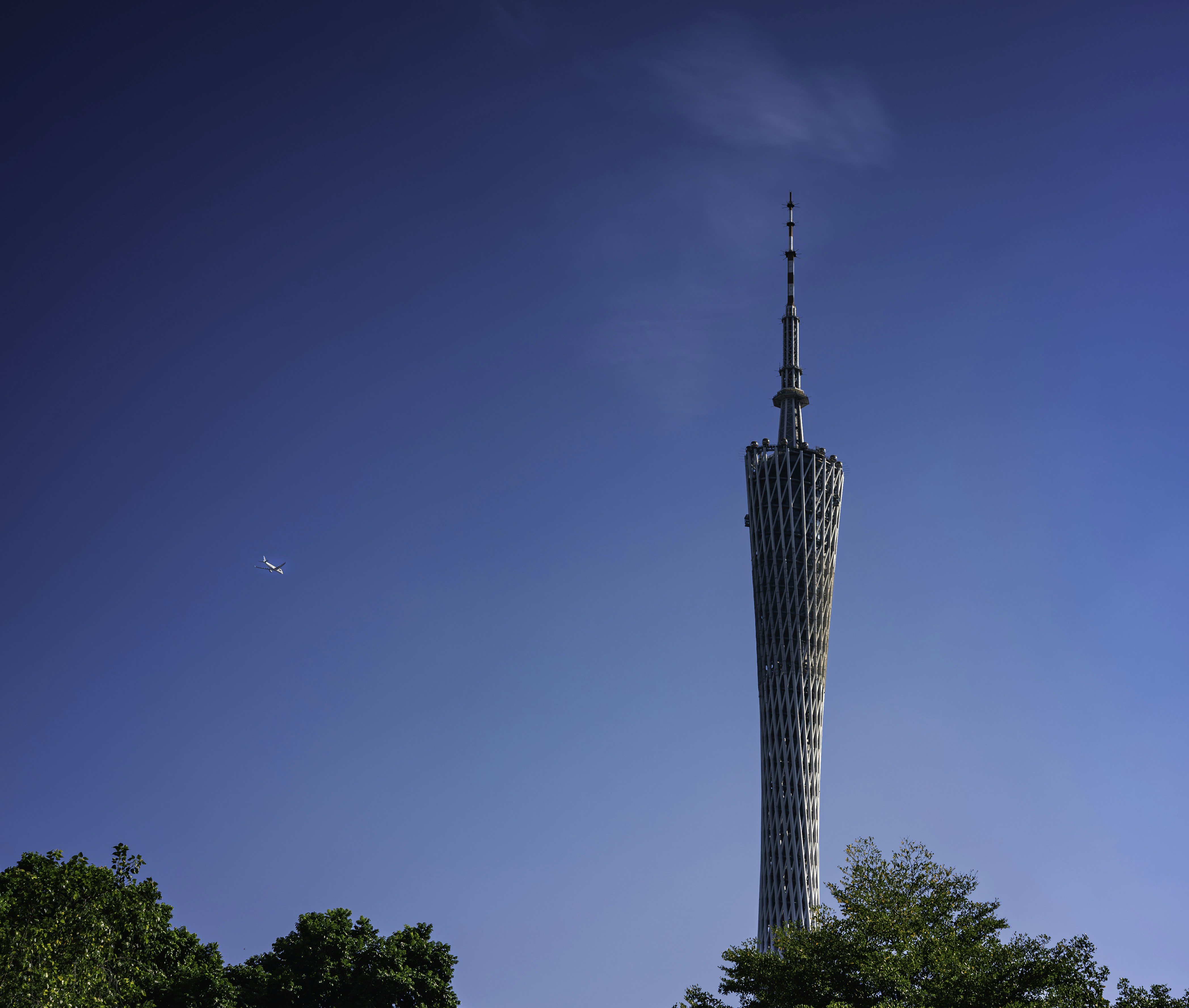 The iconic Canton Tower, with its distinctive twisting lattice structure, stands majestically against a vast, deep blue sky. Green treetops frame the lower portion of the image, and a small airplane is visible flying in the distance. This landmark is a striking feature of the Guangzhou, China skyline, captured on a bright, clear day.