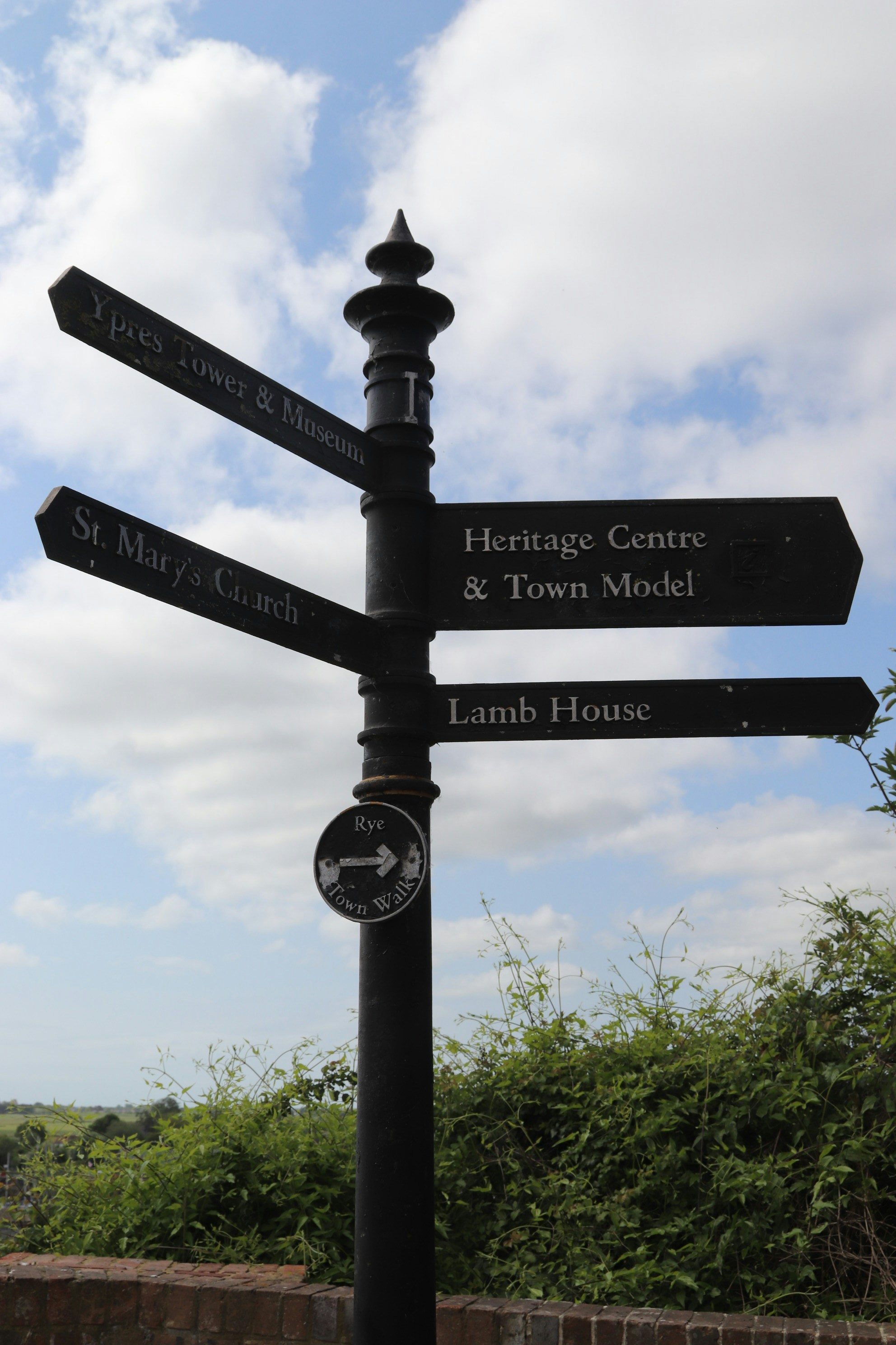 Photograph of a tall black signpost with multiple directional boards pointing to local landmarks such as Heritage Centre & Town Model, St. Mary's Church, and Lamb House, set against a bright blue sky.