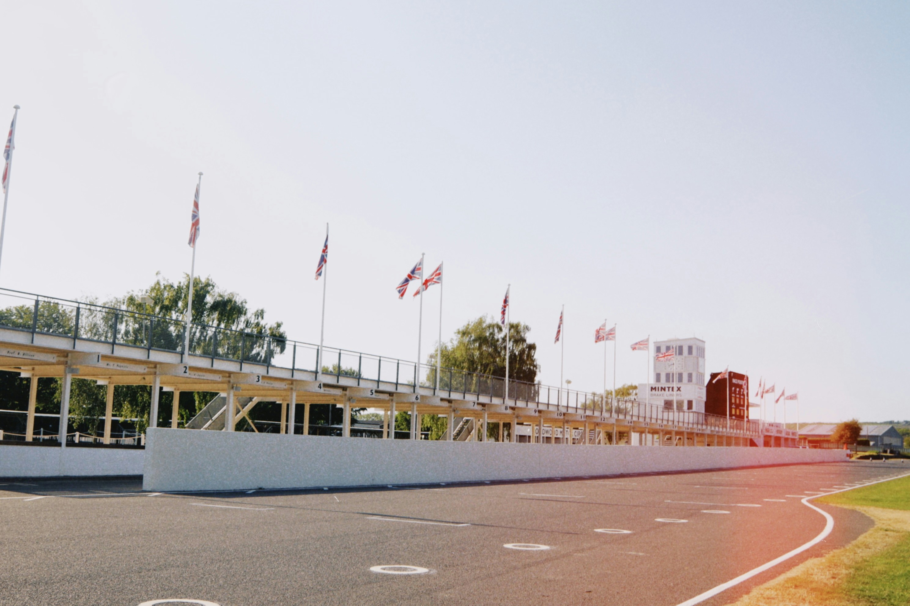Flagged Curve: Sunlit barrier and fluttering flags along a racecourse toward a distant building