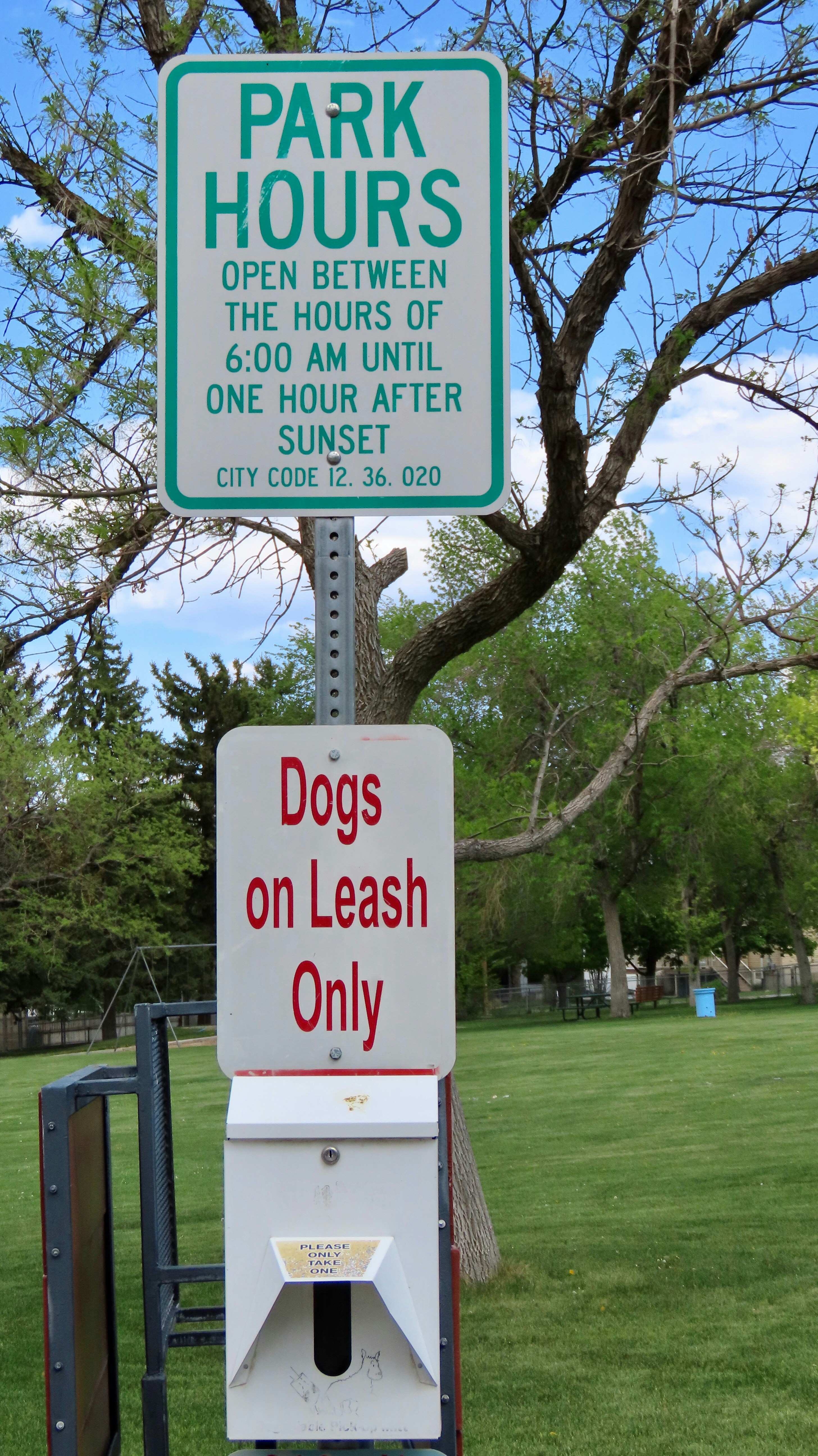 Sign detailing park hours and leash regulations for dogs, set against a backdrop of greenery and blue sky.