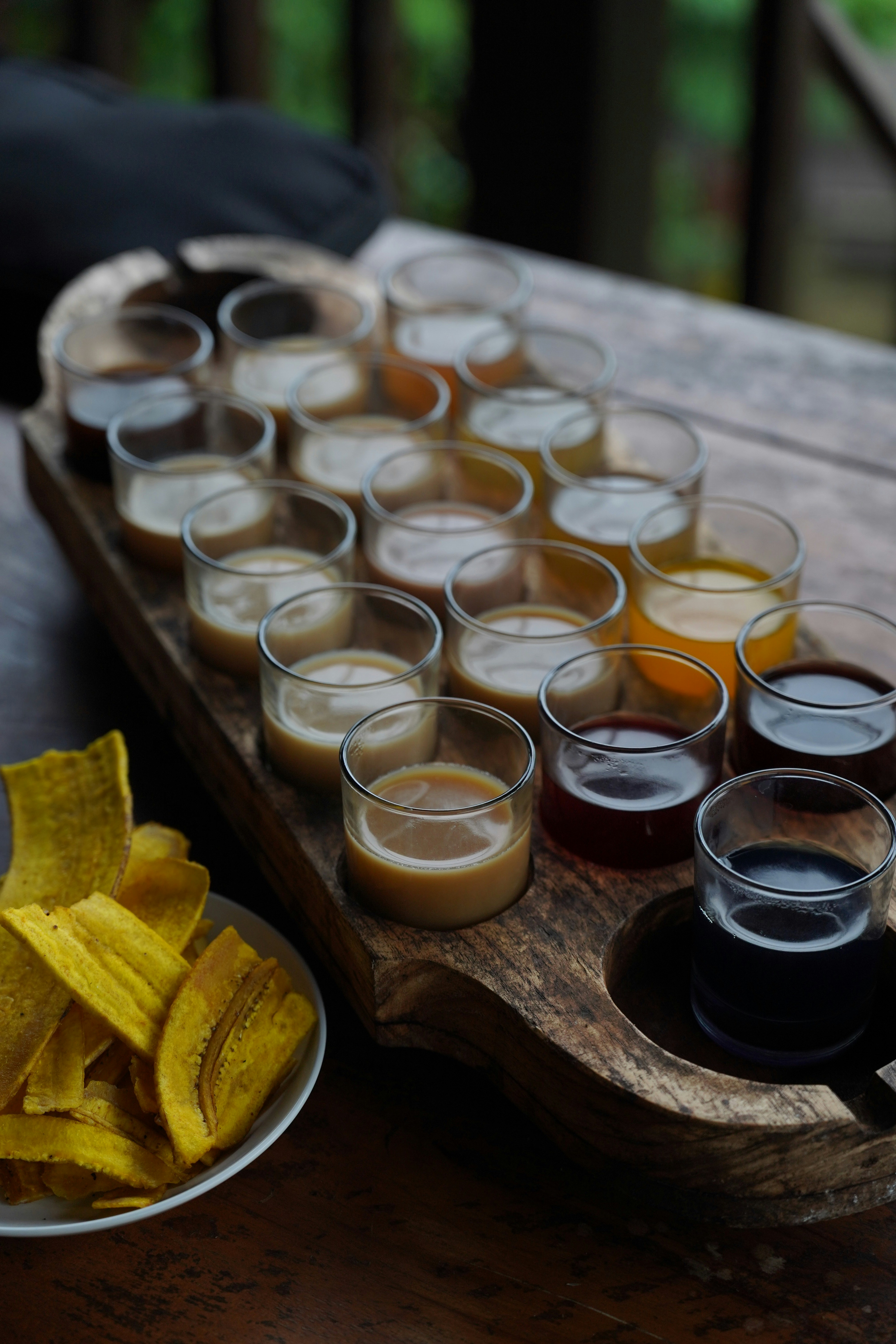 A wooden platter displays an array of colorful shot glasses filled with various beverages, accompanied by a side of crispy plantain chips.