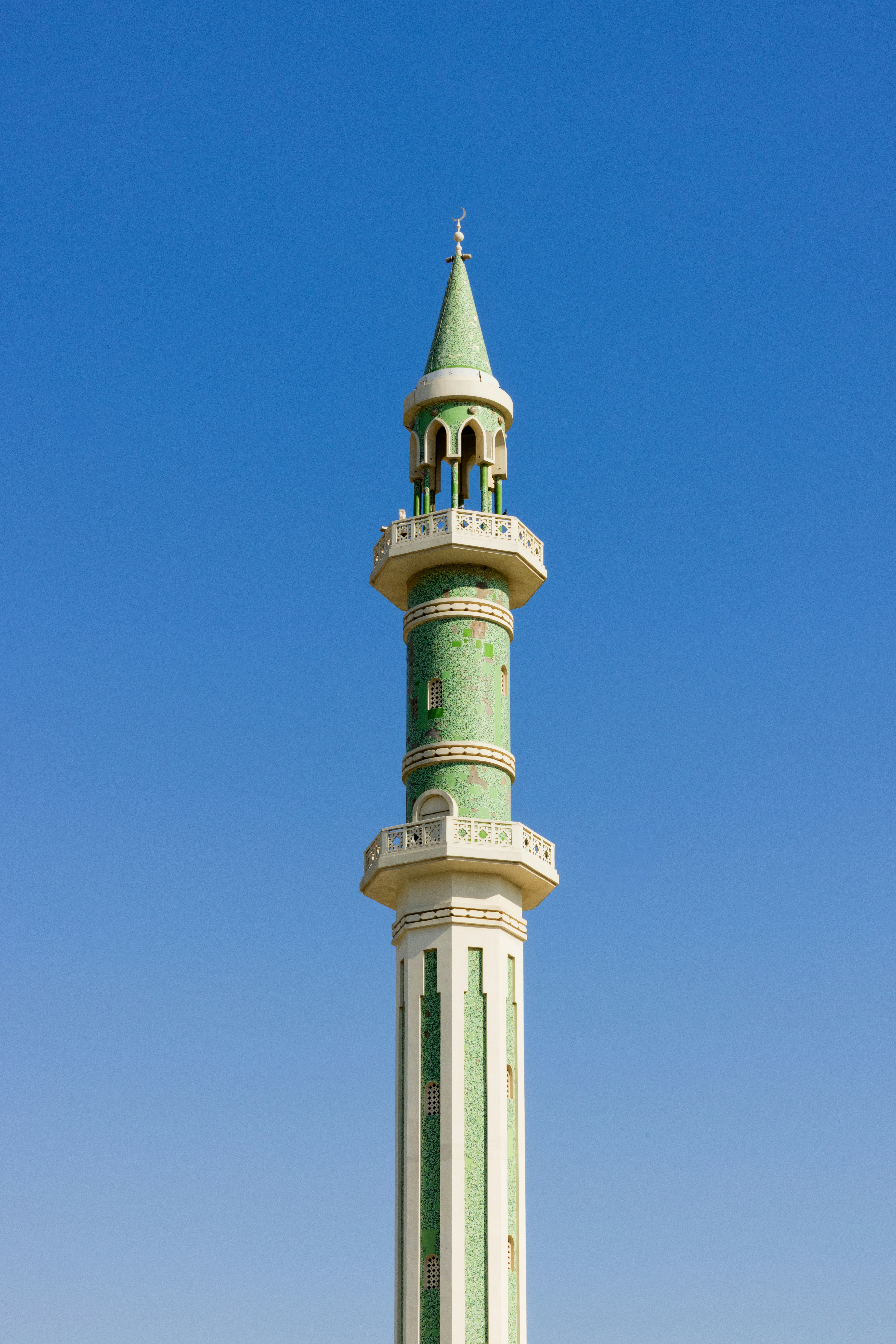 Tall green and white minaret reaching towards a clear blue sky, showcasing intricate architectural details. The structure stands as a prominent feature against the backdrop of the horizon.