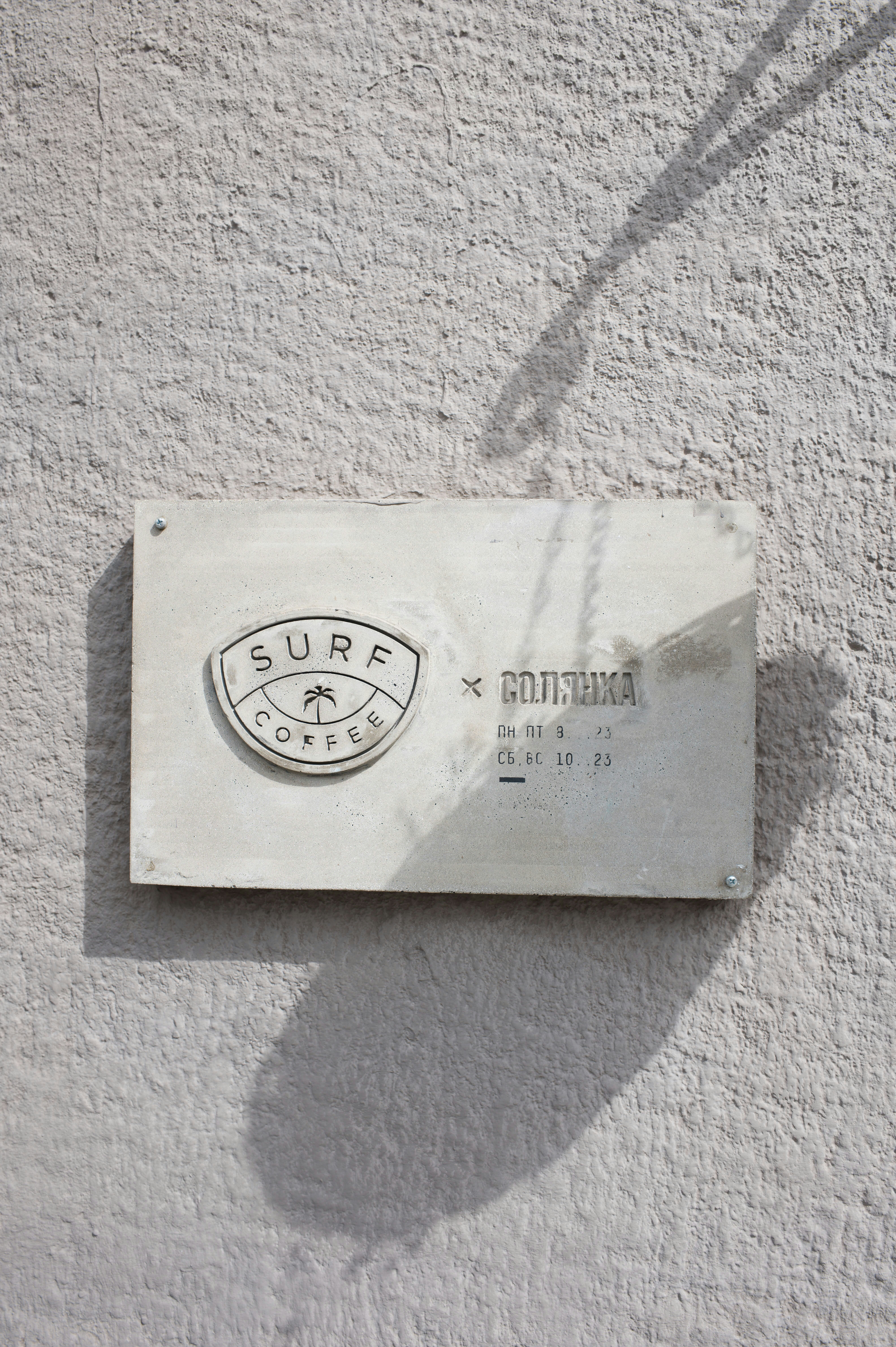 Minimalist scene featuring a metal plaque mounted on a pale stucco wall, with a pronounced shadow arc crossing above and to the right of the plaque.