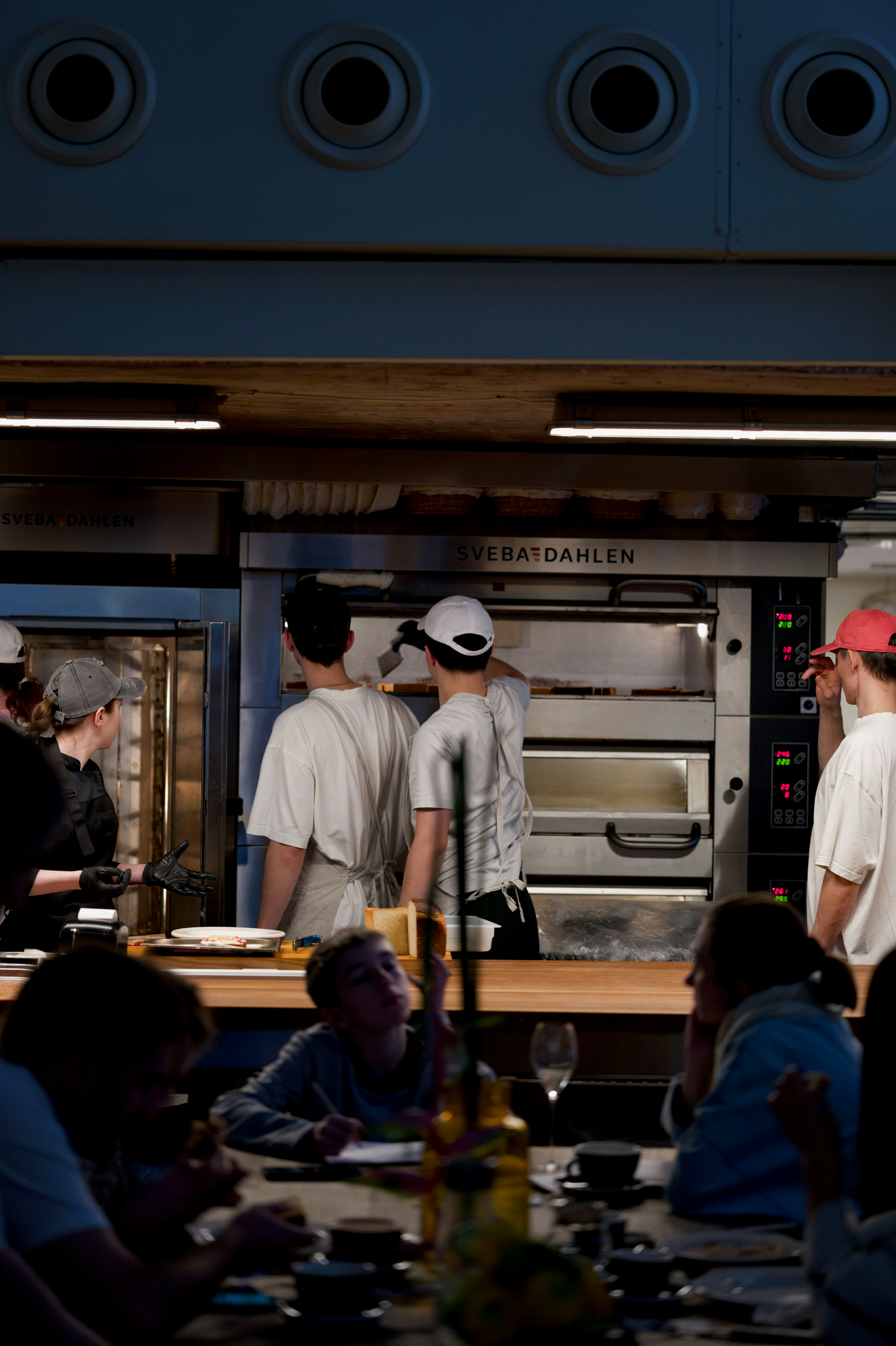 Chefs collaborate in a bustling kitchen, focused on their culinary creations while diners enjoy the atmosphere nearby.