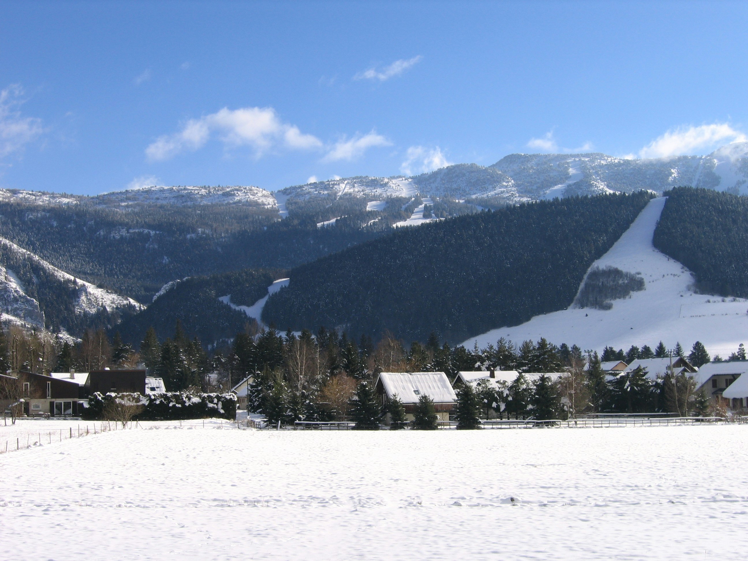 A picturesque winter landscape featuring snow-laden mountains and quaint houses nestled in a snowy field under a bright blue sky.