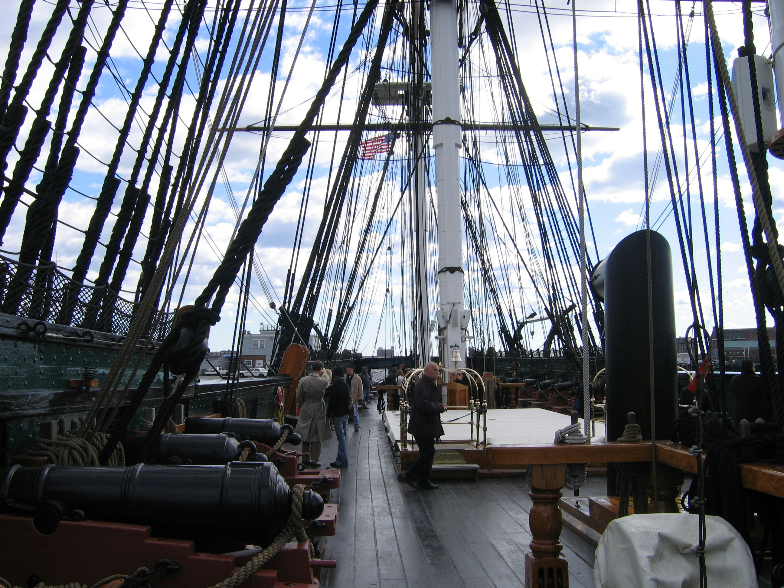 Visitors explore the deck of a historic ship, surrounded by masts and rigging against a cloudy sky.