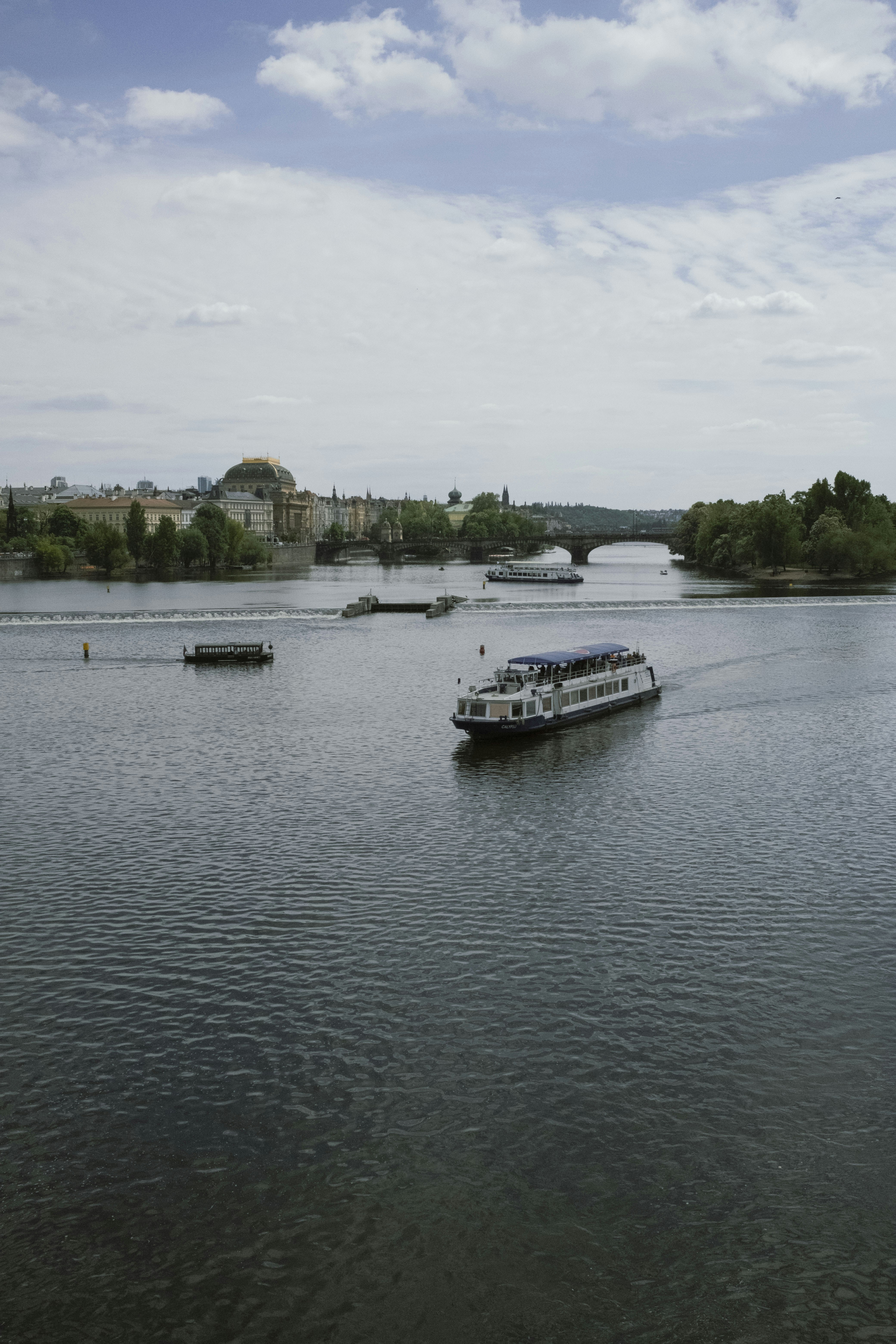 A riverboat gliding through the calm waters of the Vltava River, framed by lush greenery and historic architecture. The scene captures the tranquil essence of leisurely river travel.