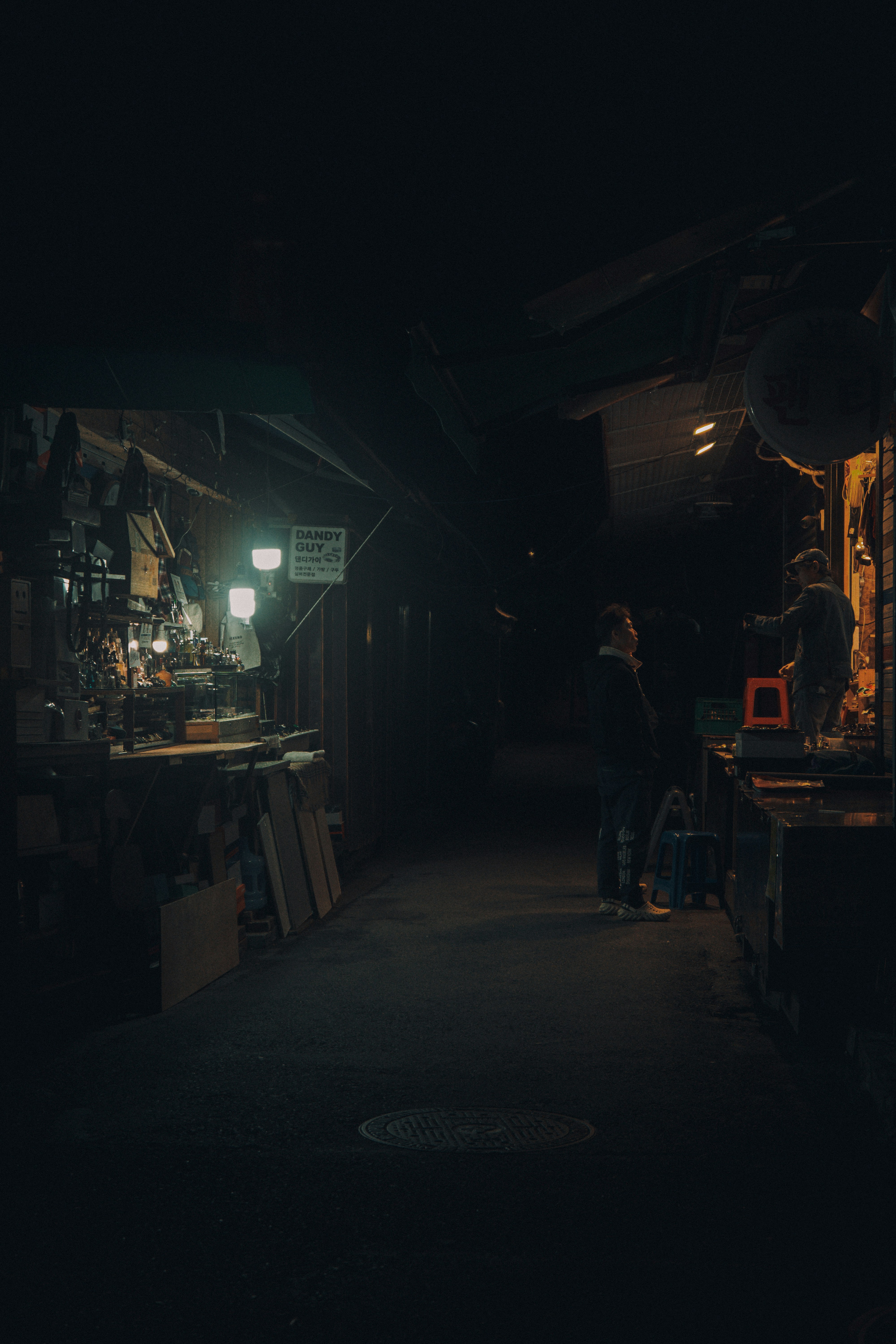 Dimly lit alleyway showcasing a bustling night market with vendors and patrons. A lone figure stands in contemplation amidst the vibrant stalls.
