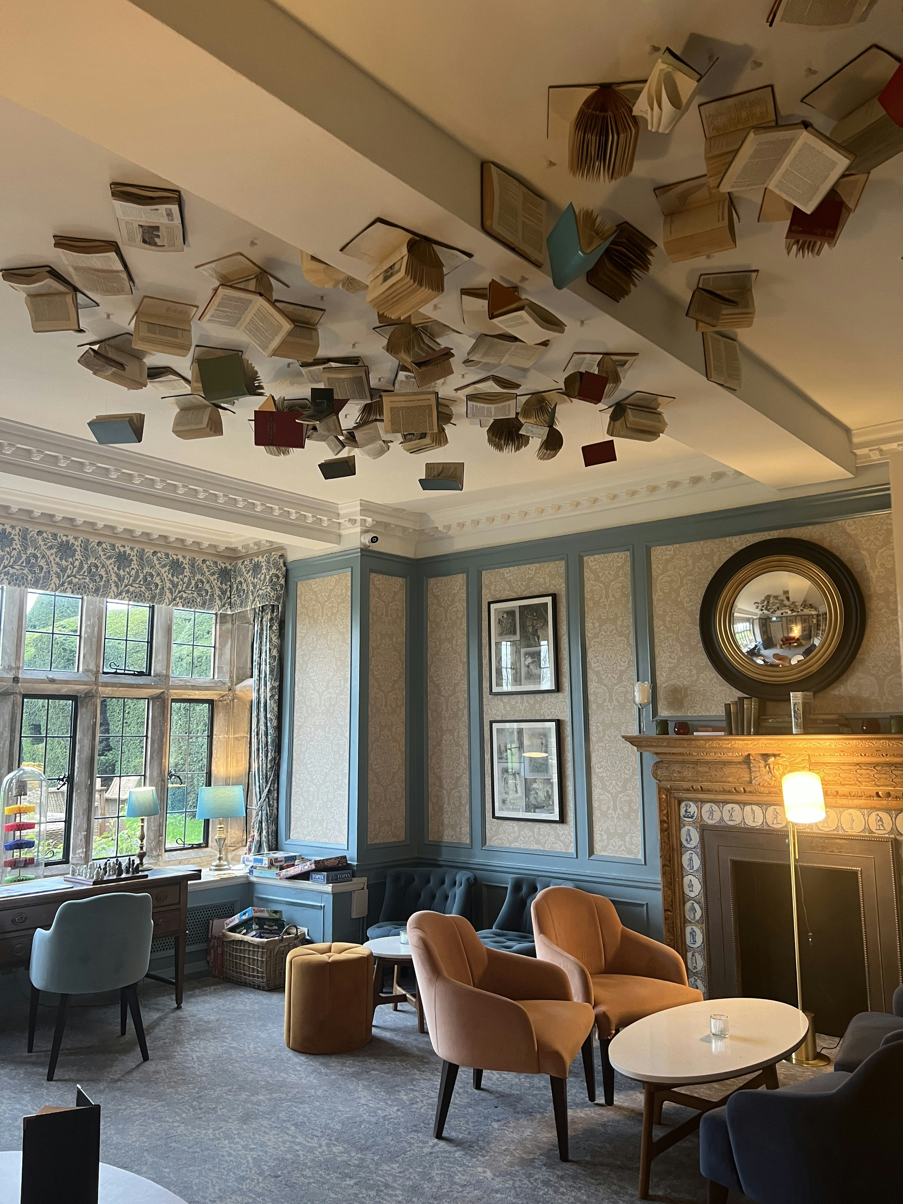 Cozy parlour with blue paneling, mid-century chairs, and a dramatic ceiling of floating books; a literary art installation high above an intimate living space.