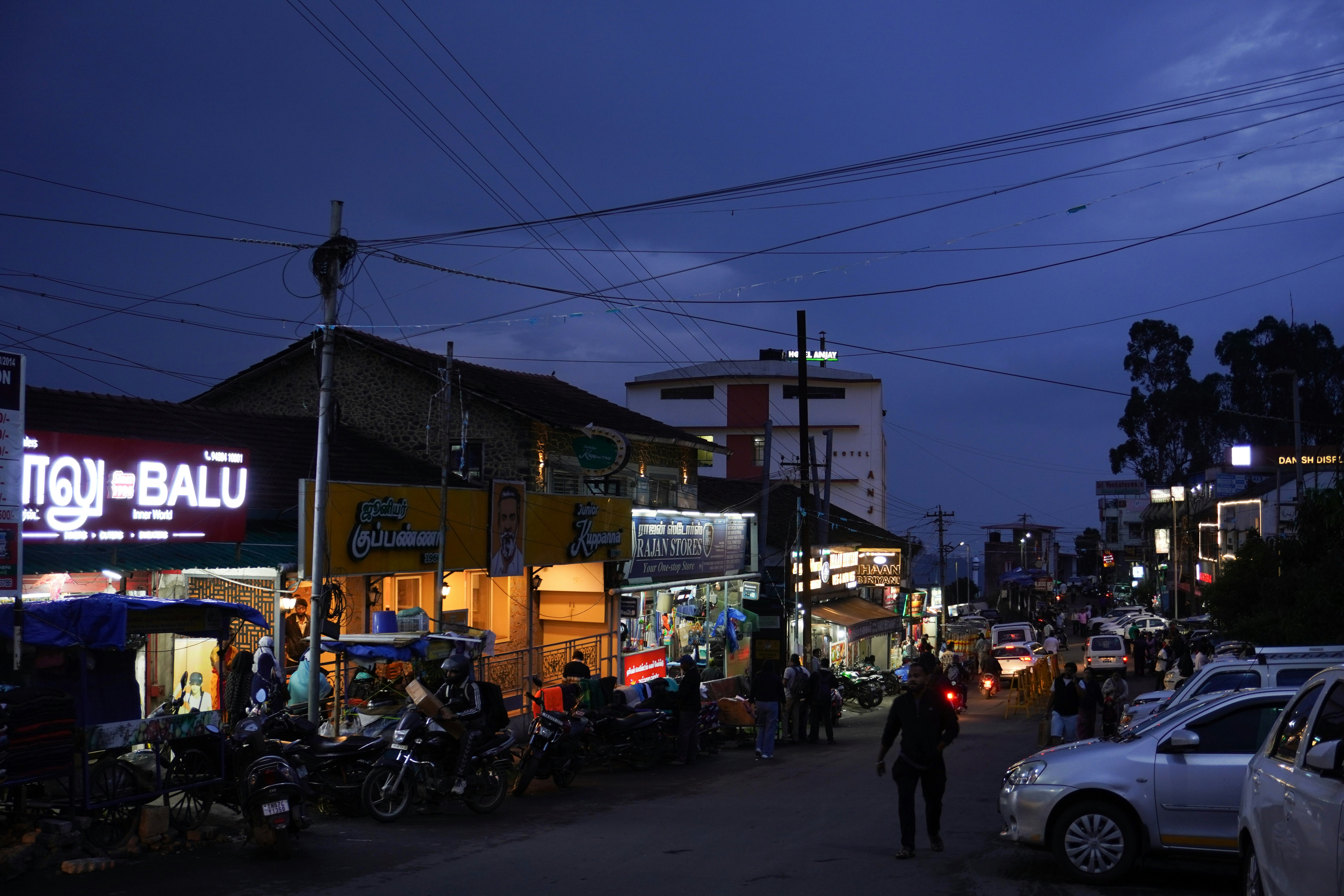 Vibrant street scene at dusk featuring illuminated shop signs and bustling activity among pedestrians and vehicles. The atmosphere reflects the lively essence of urban commerce.