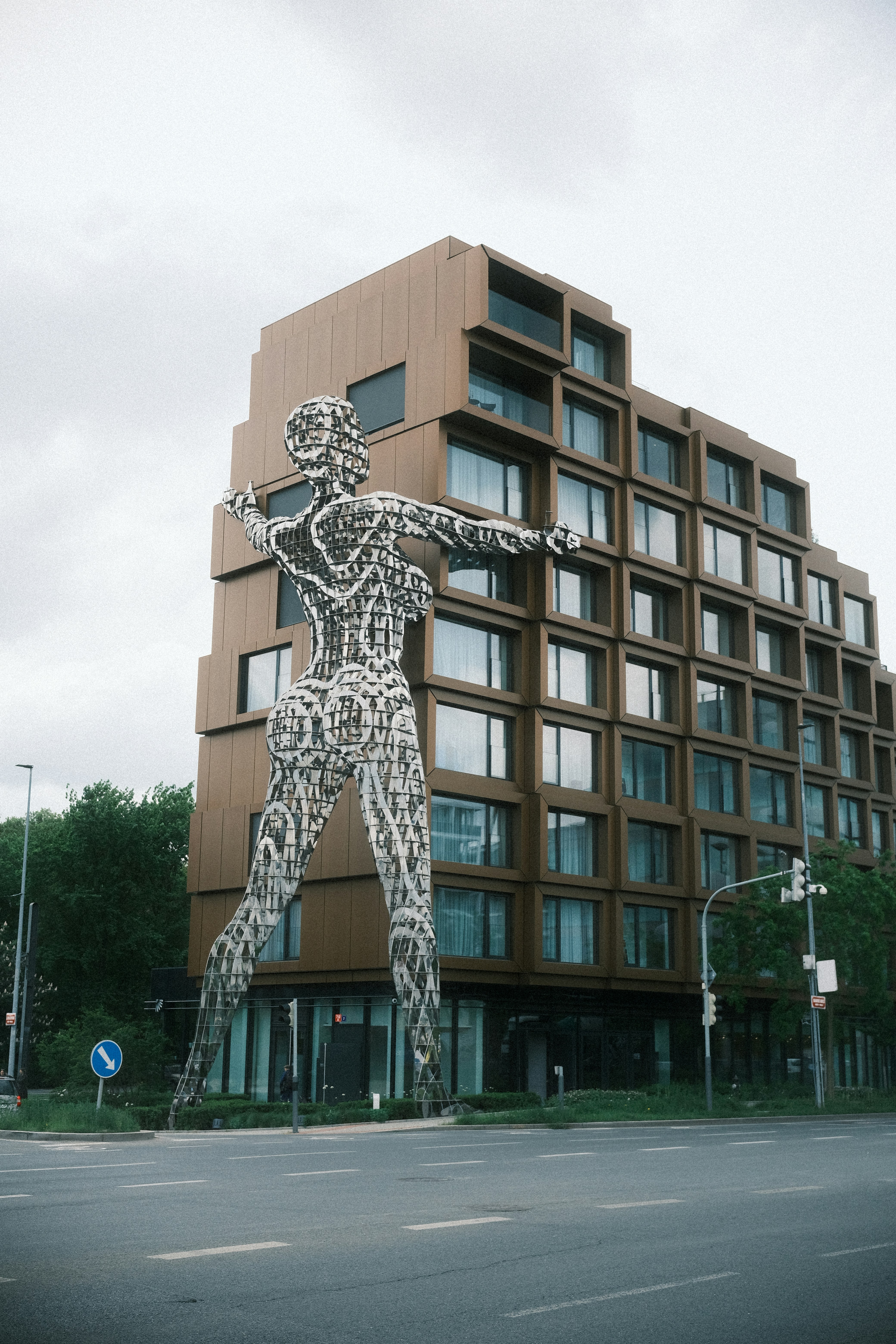 A large, striking sculpture of a figure made of dark material contrasts with a contemporary building. The setting suggests an artistic urban area on a cloudy day.