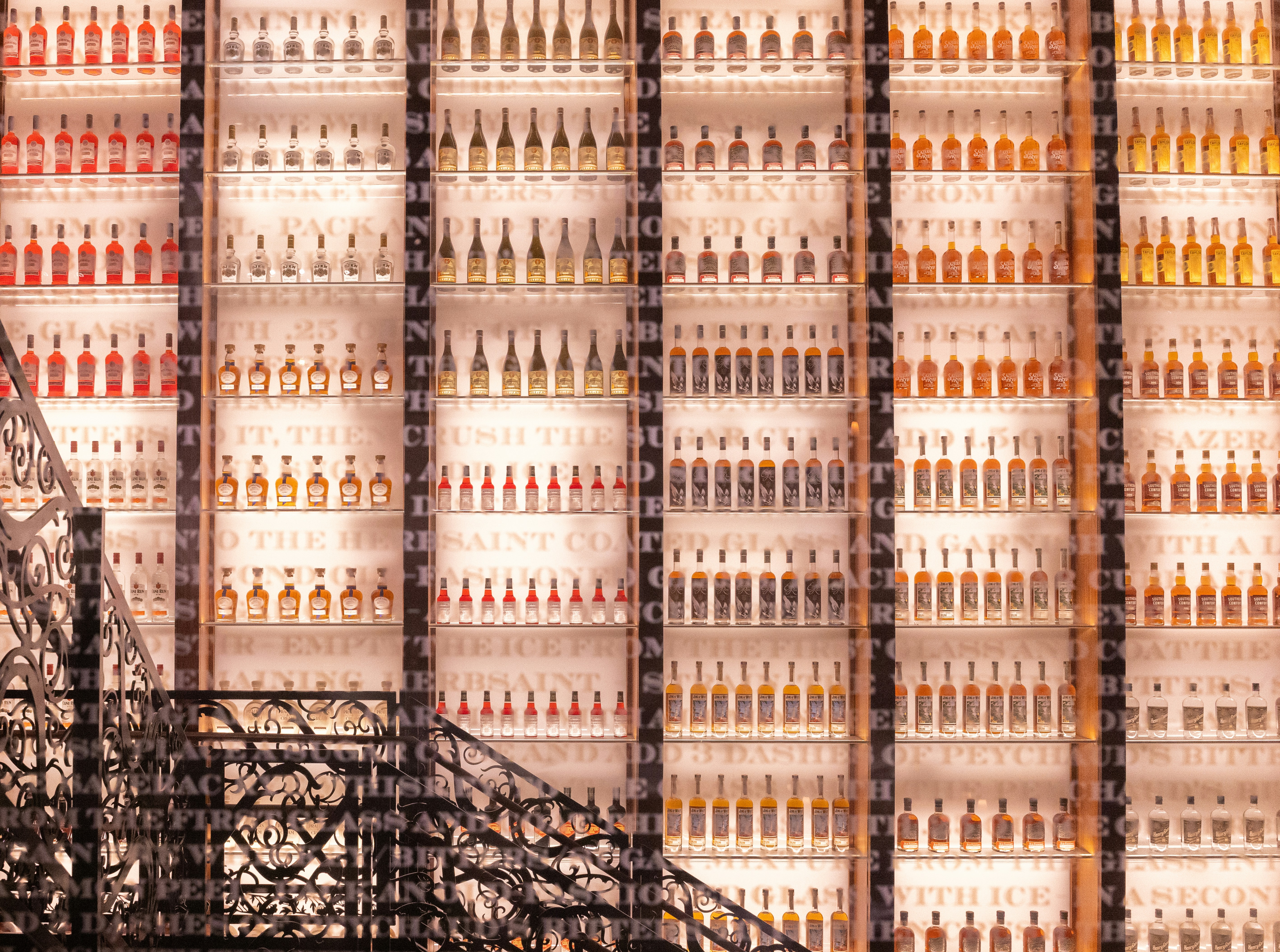 A meticulously arranged display of various liquor bottles on illuminated shelves, showcasing a spectrum of colors and designs. Elegant wrought iron staircase adds depth to the scene.