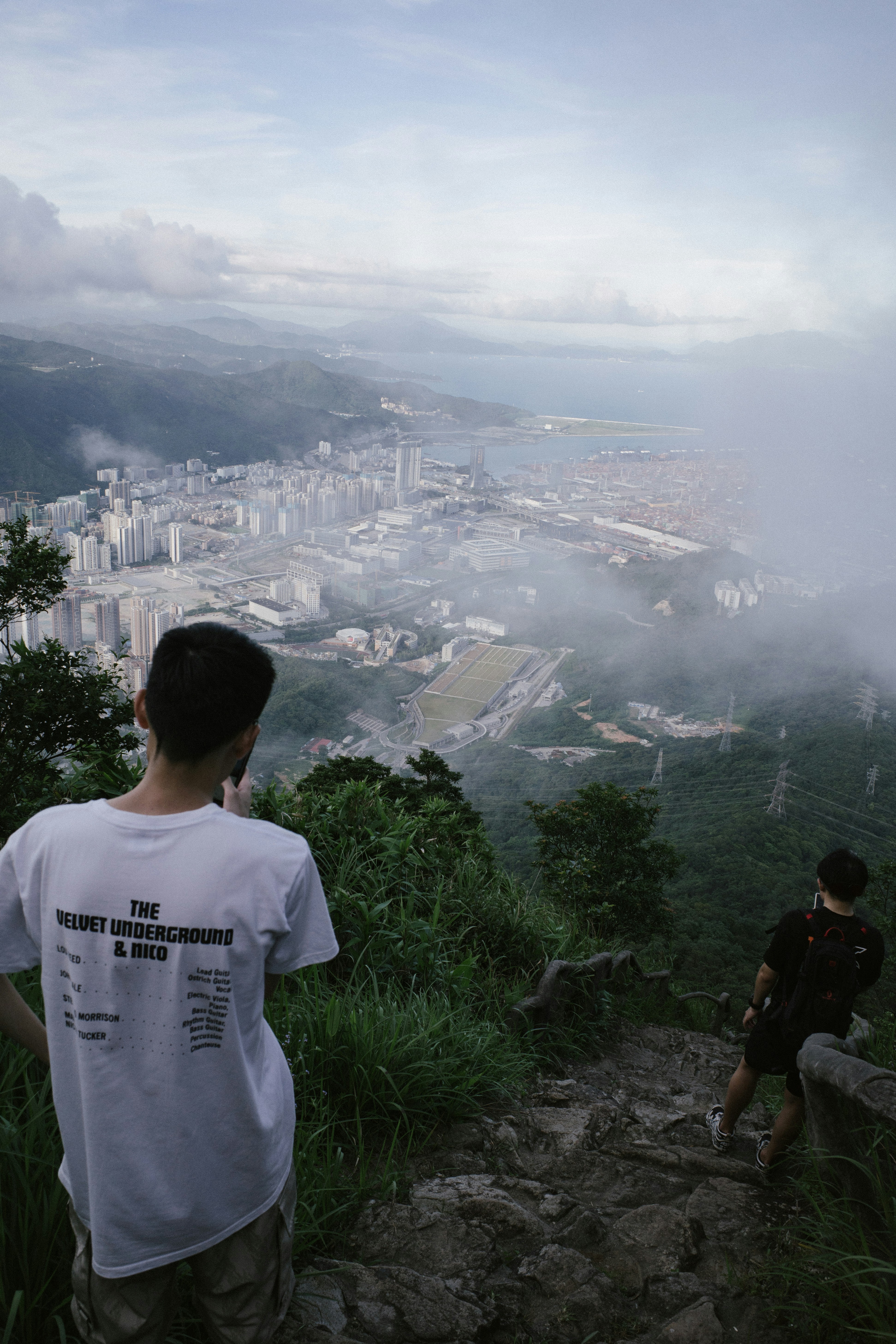 Hikers overlooking a sprawling cityscape surrounded by mountains and mist. The scene captures the contrast between nature and urban development.