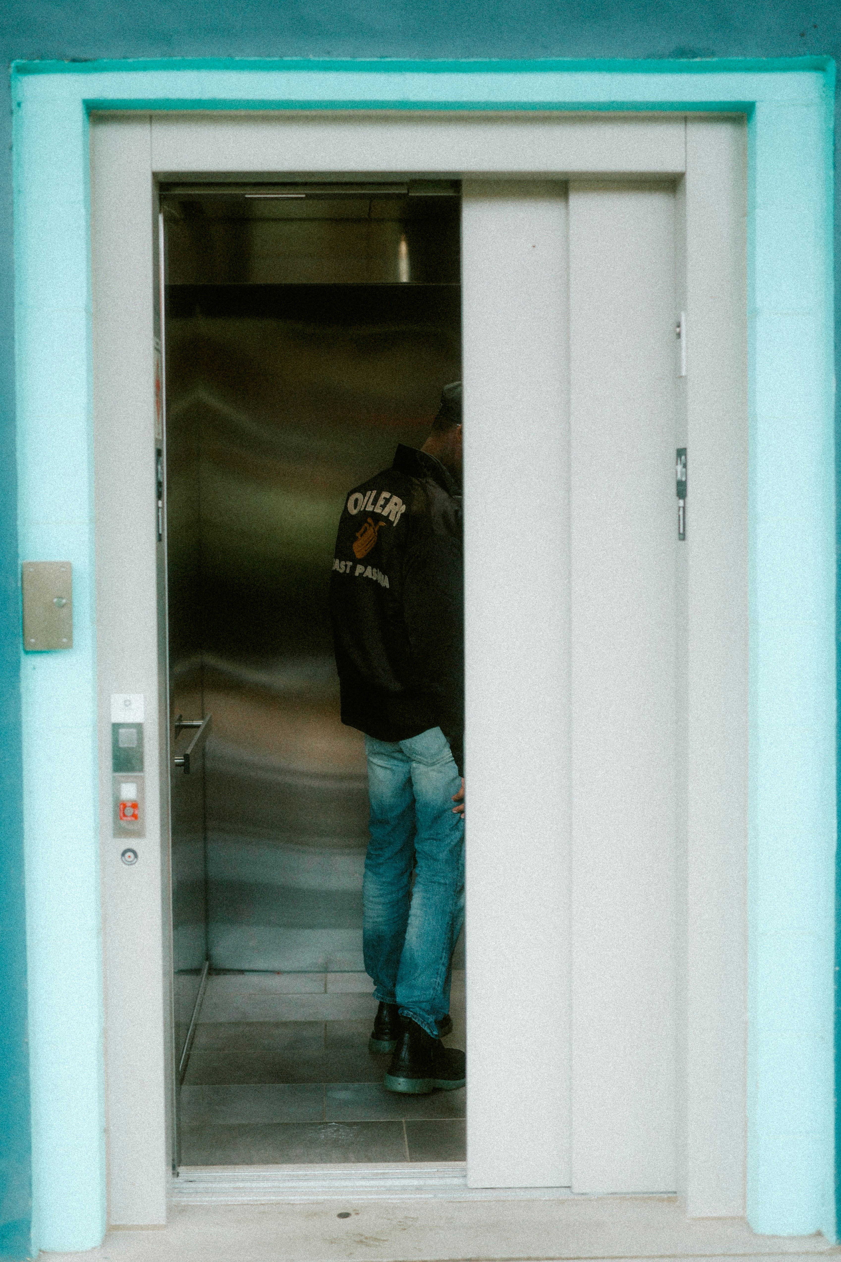 Turquoise Portal: A Back-to-Camera Figure in a Narrow Elevator Frame