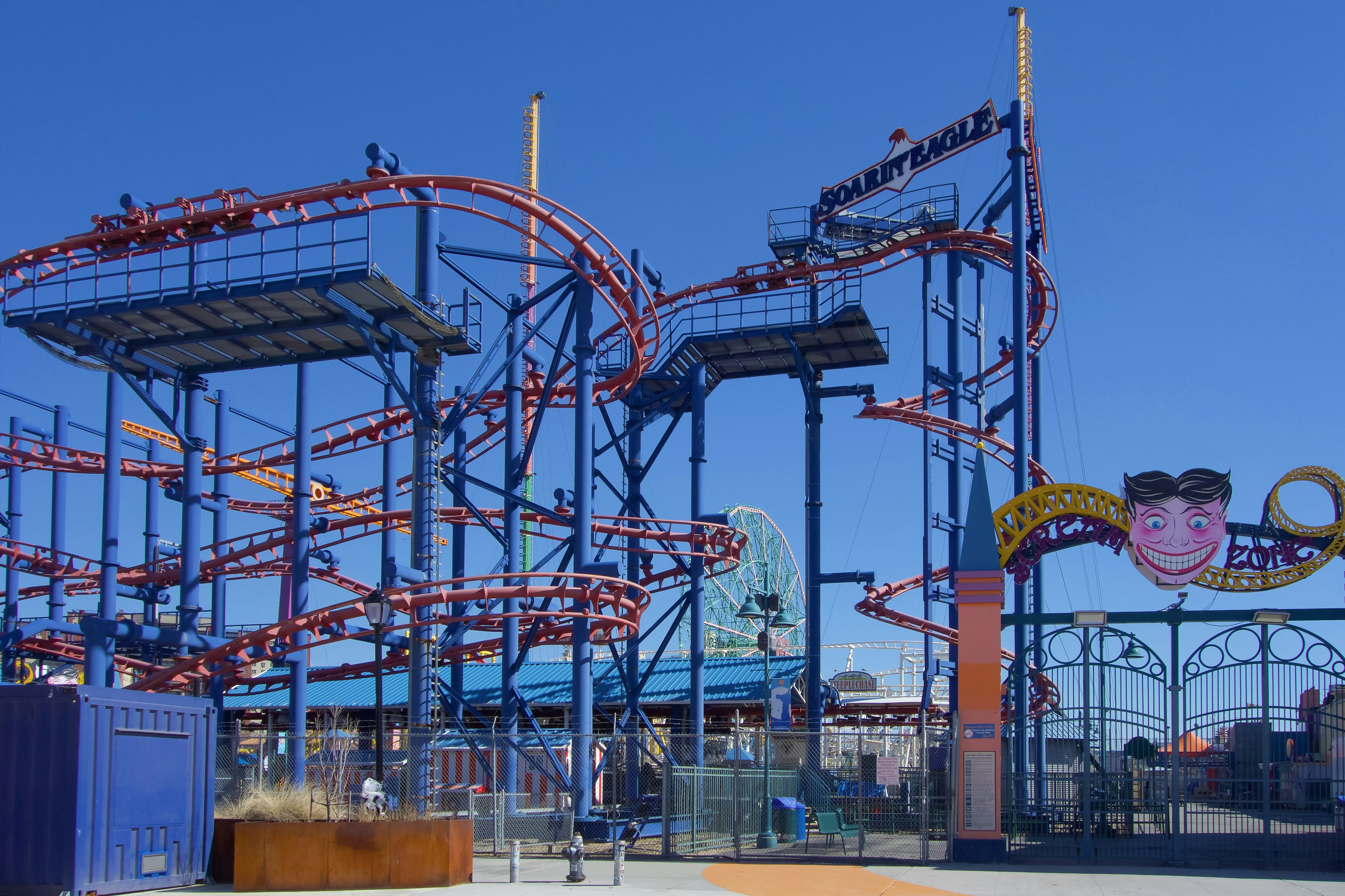 Blue Steel Lattice of Motion: Sunlit Coaster Arcs Over a Quiet Amusement Entrance