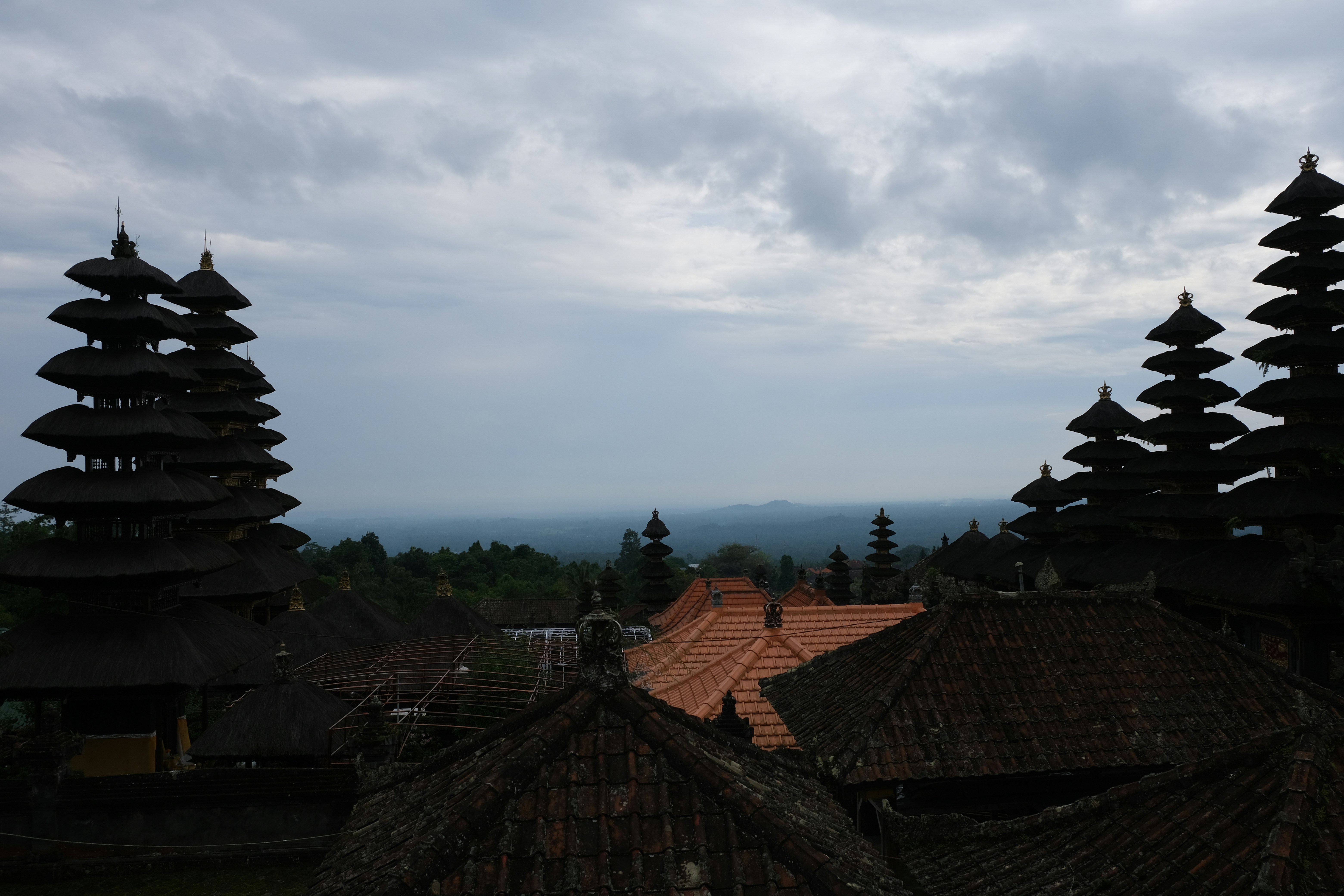 Majestic Balinese temples rise against a moody sky, showcasing intricate architecture and terracotta roofs. A serene landscape stretches into the distance.