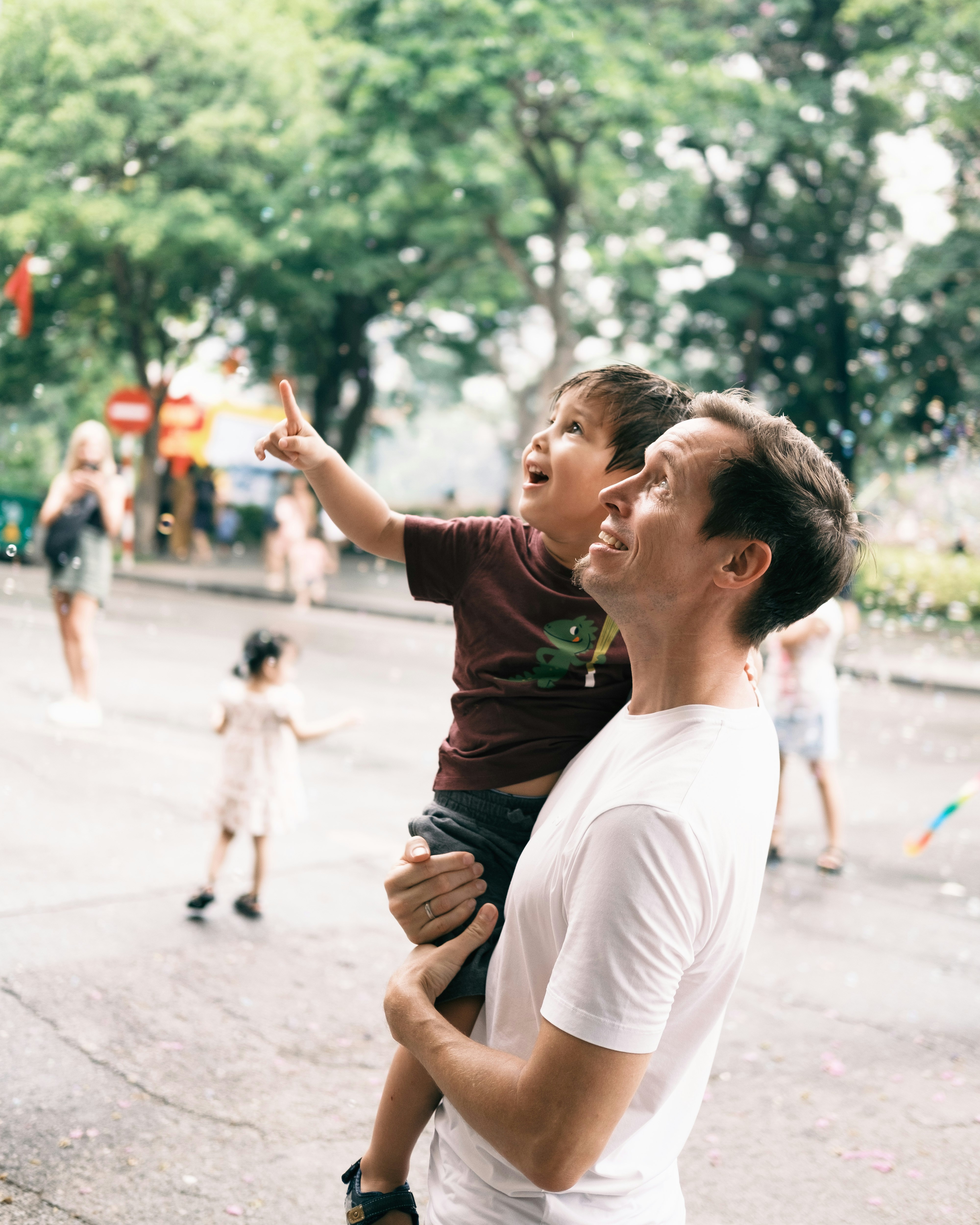 Upward Wonder: A Father Lifts His Son Amid a Confetti-Sprinkled Street Moment