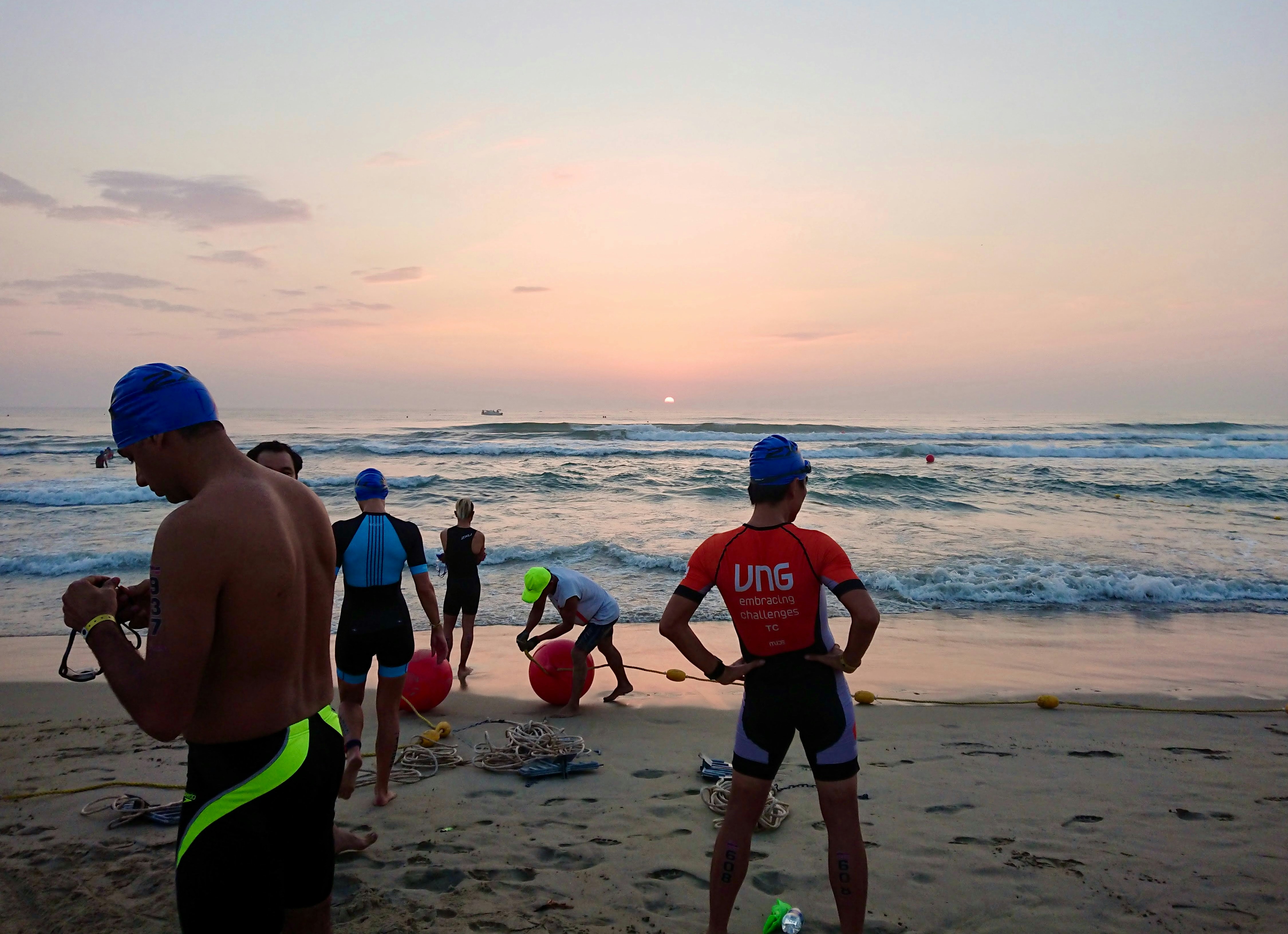 Triathletes in swim caps gather on the beach as the sun sets over the horizon, preparing for an upcoming competition.