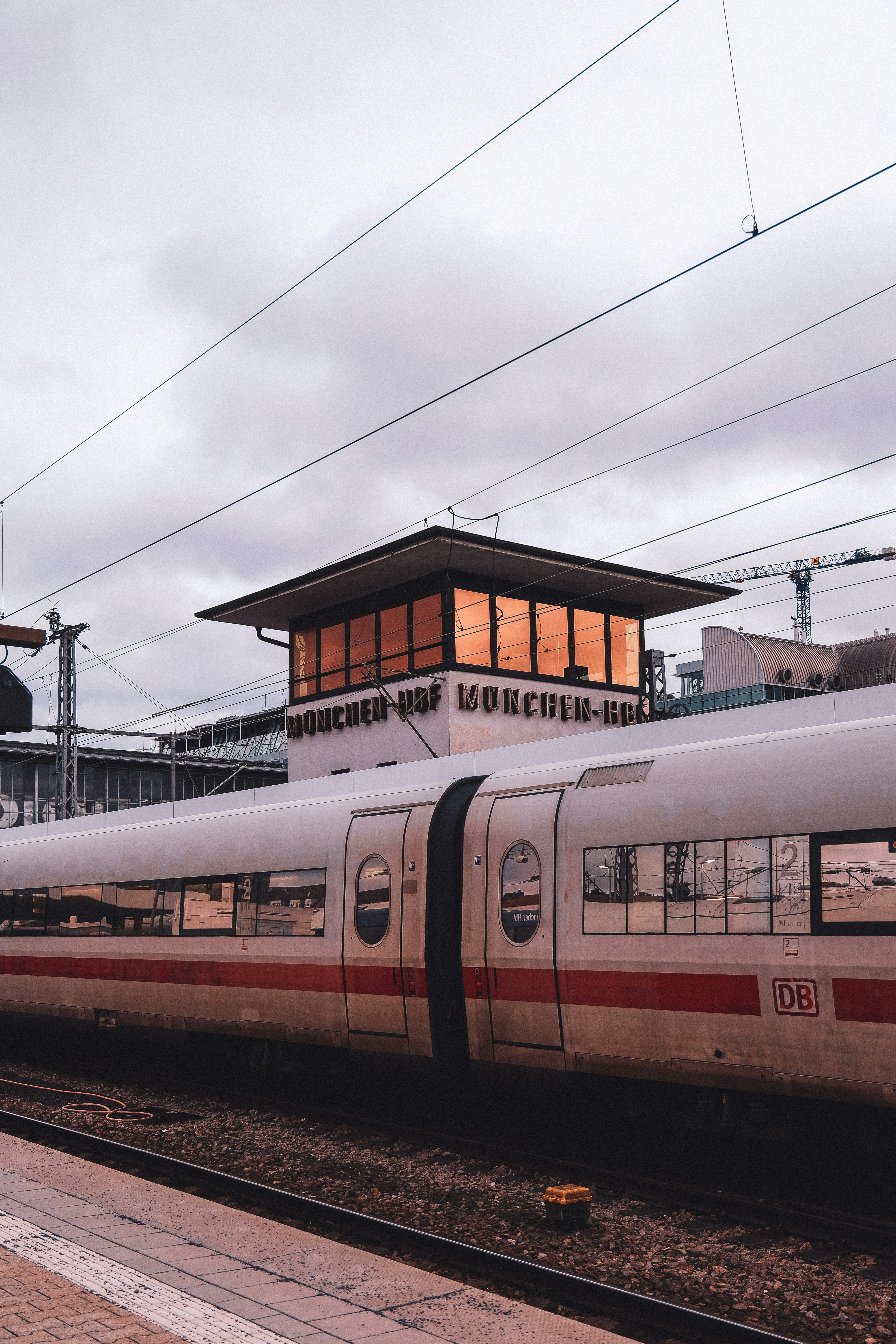 An ICE high-speed train at Munich Central Station (München Hbf) under a cloudy sky. The control tower's glowing windows add warmth to the industrial atmosphere of the scene.