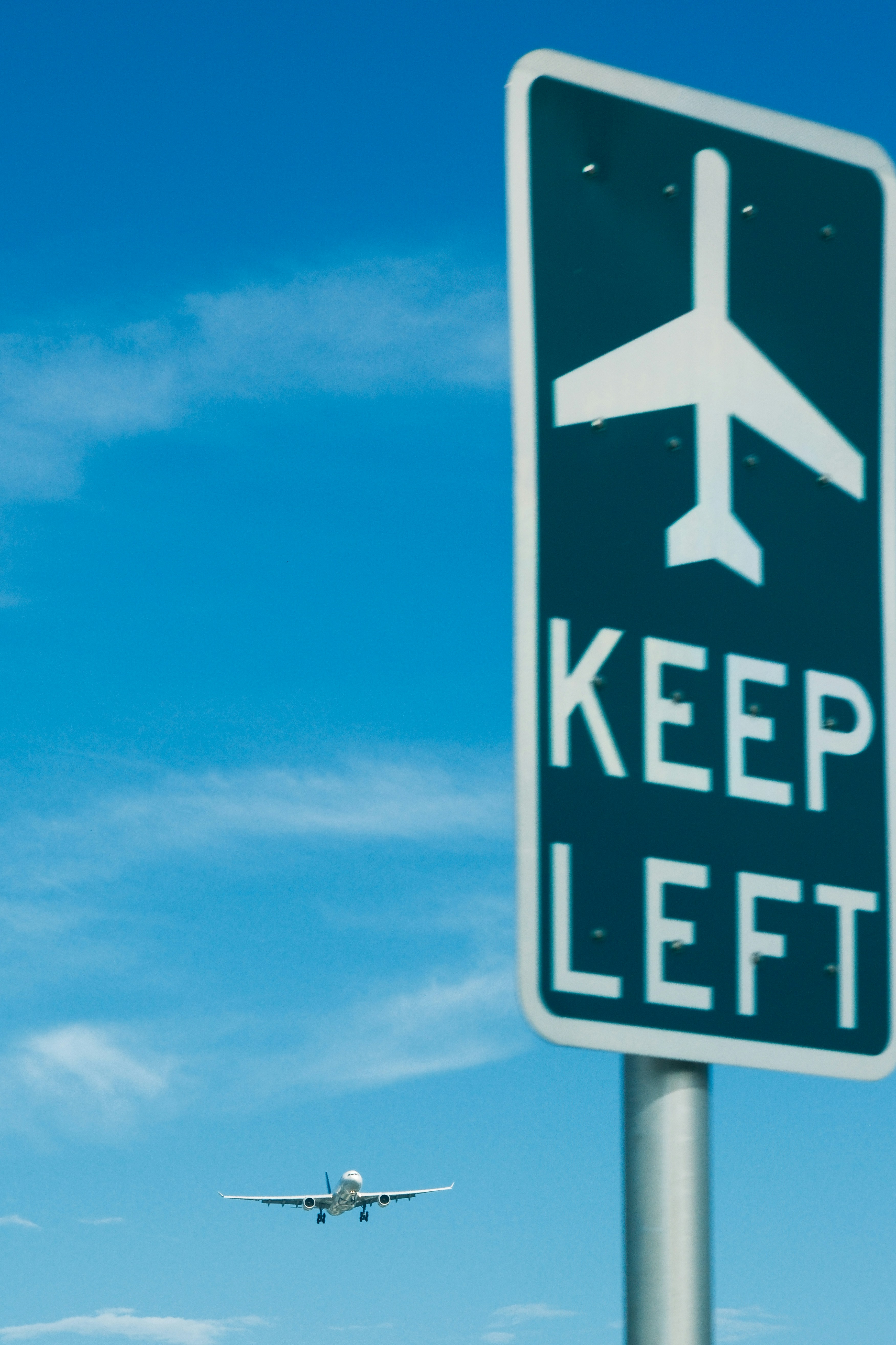 A landing plane in the distance and a bold airport sign in the foreground share the blue sky in this layered travel scene.