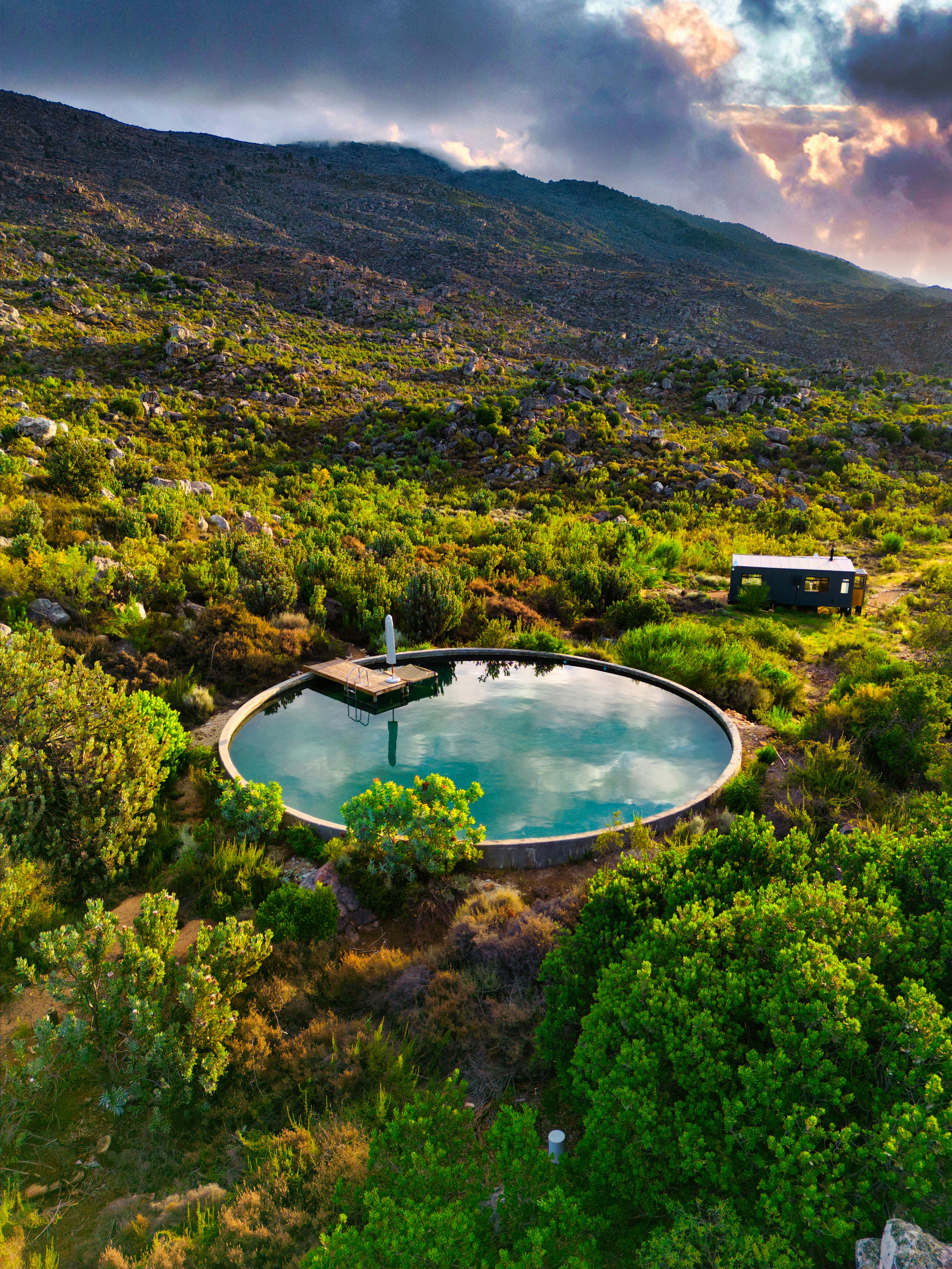 Circular pool surrounded by lush greenery and rocky terrain under a dramatic sky.