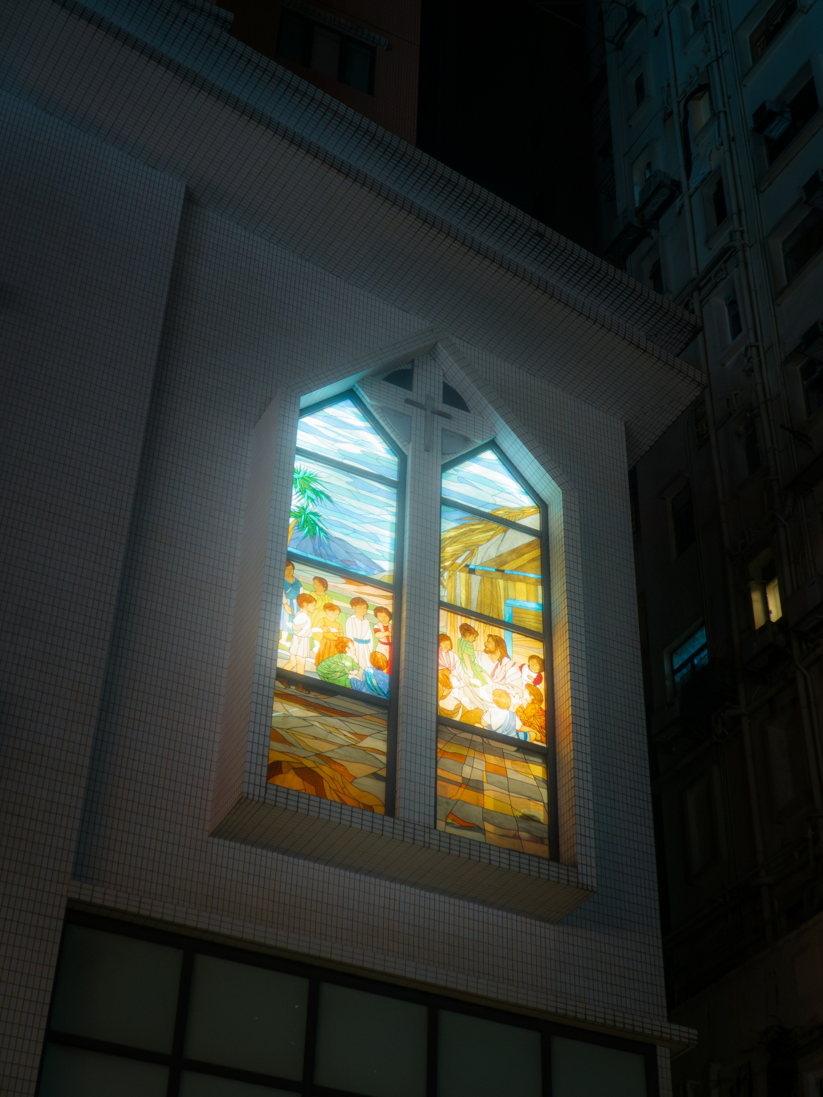 The exterior wall of a church building in Mong Kok, one of the busiest districts in Hong Kong, stands out among old buildings. A large, colorful stained glass window, backlit at night, depicts a religious scene with Jesus and several children. The vibrant colors glow warmly against the dark surroundings, creating a striking focal point in the bustling urban landscape.
