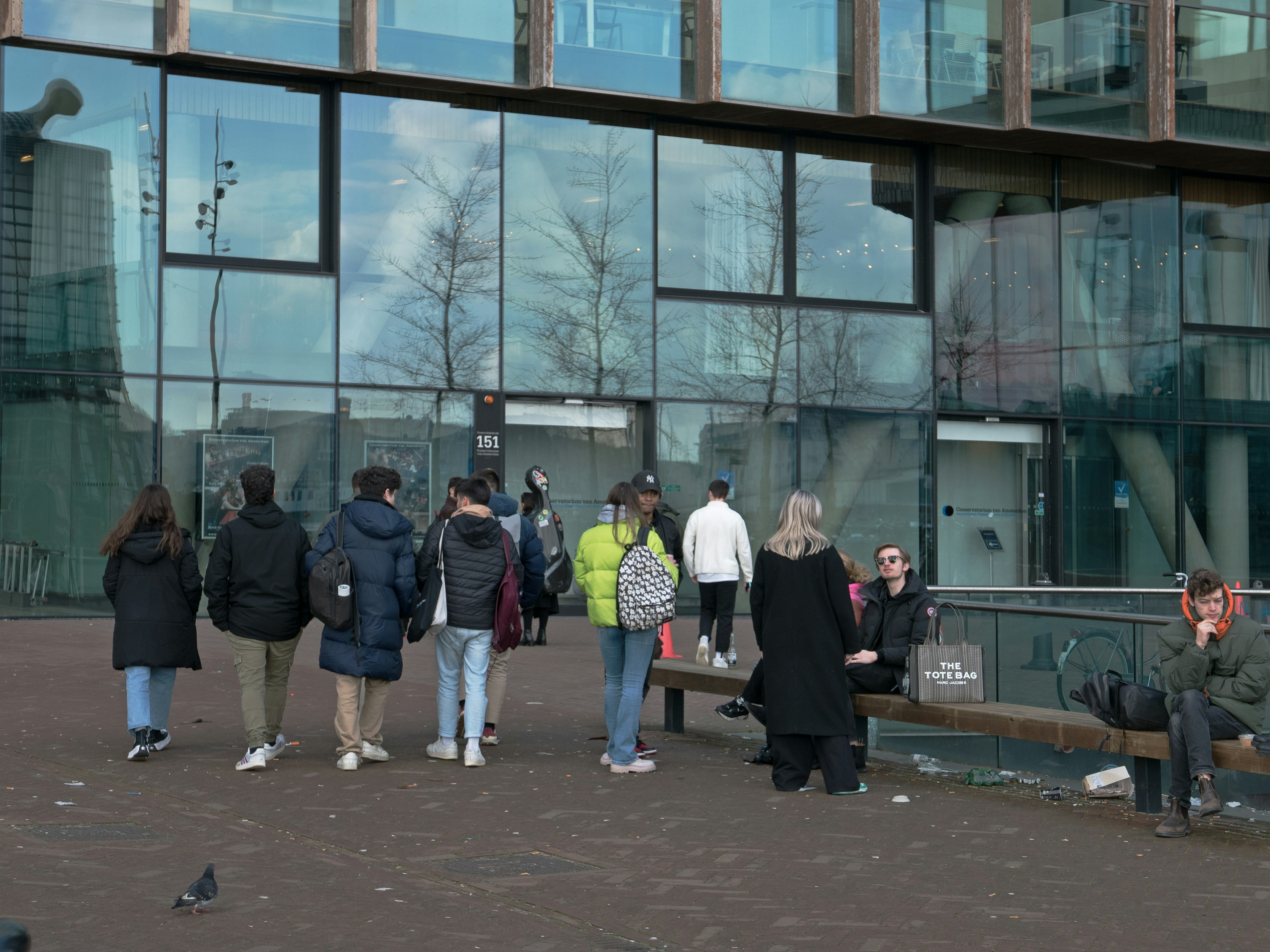 Free photo of students, waiting in front of the large glass window facade of the conservatory building - modern glass architecture with the interior of pile constructions. I like the transparency and the reflected light. Street photography of The Netherlands in free download images by Dutch photographer Fons Heijnsbroek, March 2024. | People stand outside a modern glass building.