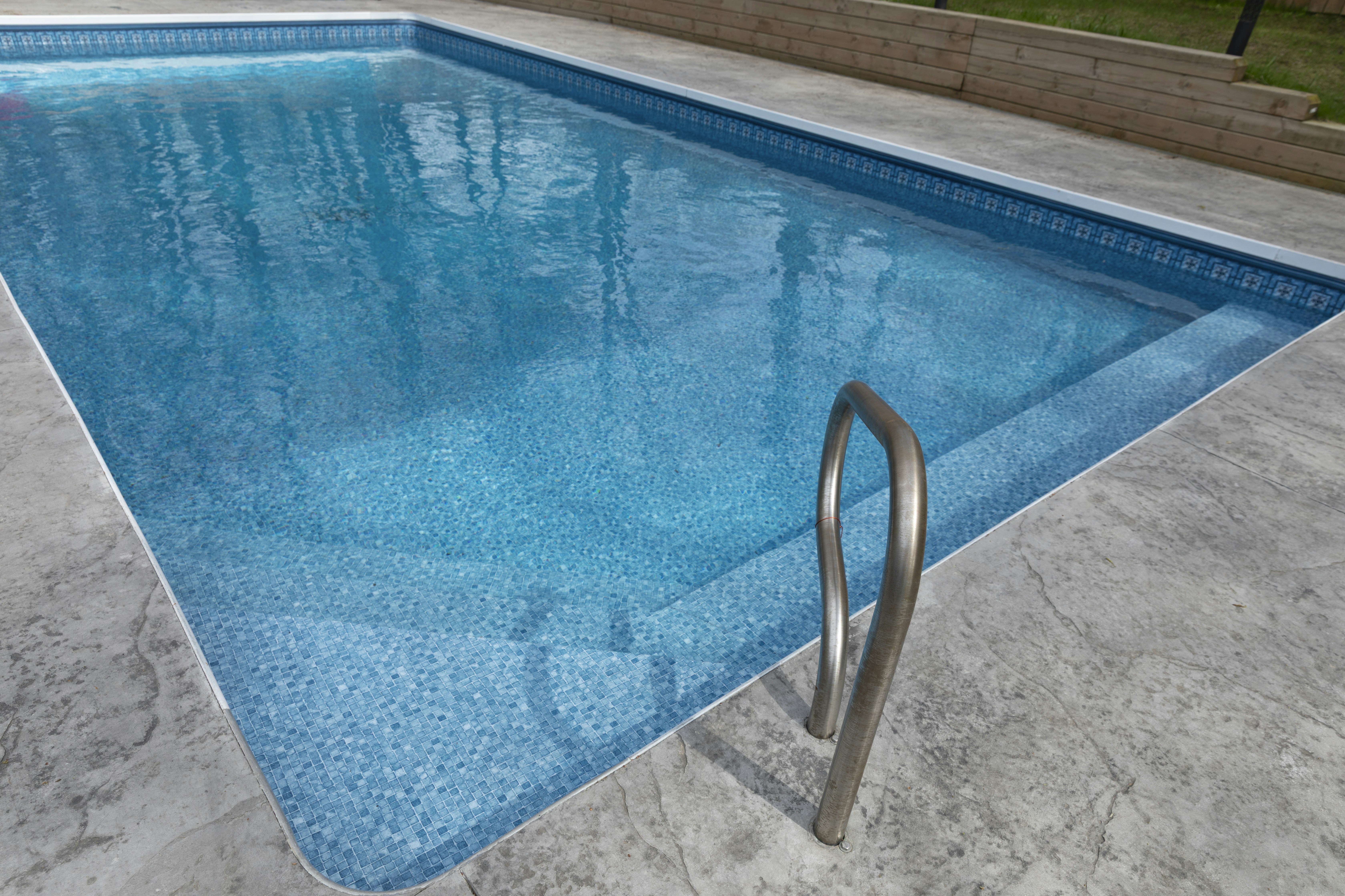 Crystal-clear swimming pool reflecting the sky, with a sleek metallic ladder leading into the water. The surrounding area features a textured concrete deck.