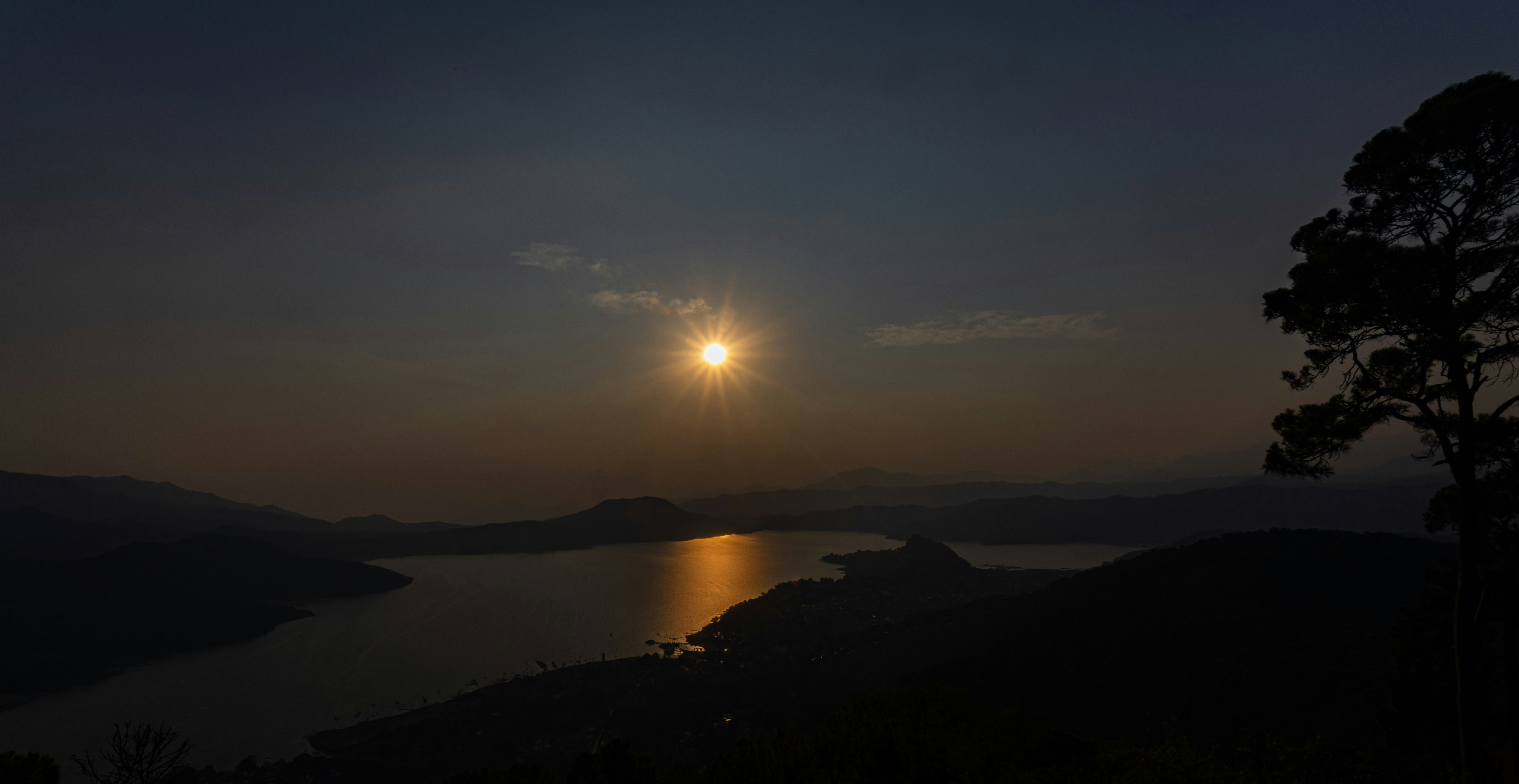 Golden sun setting over a serene lake, casting reflections on still water with distant hills framing the horizon.