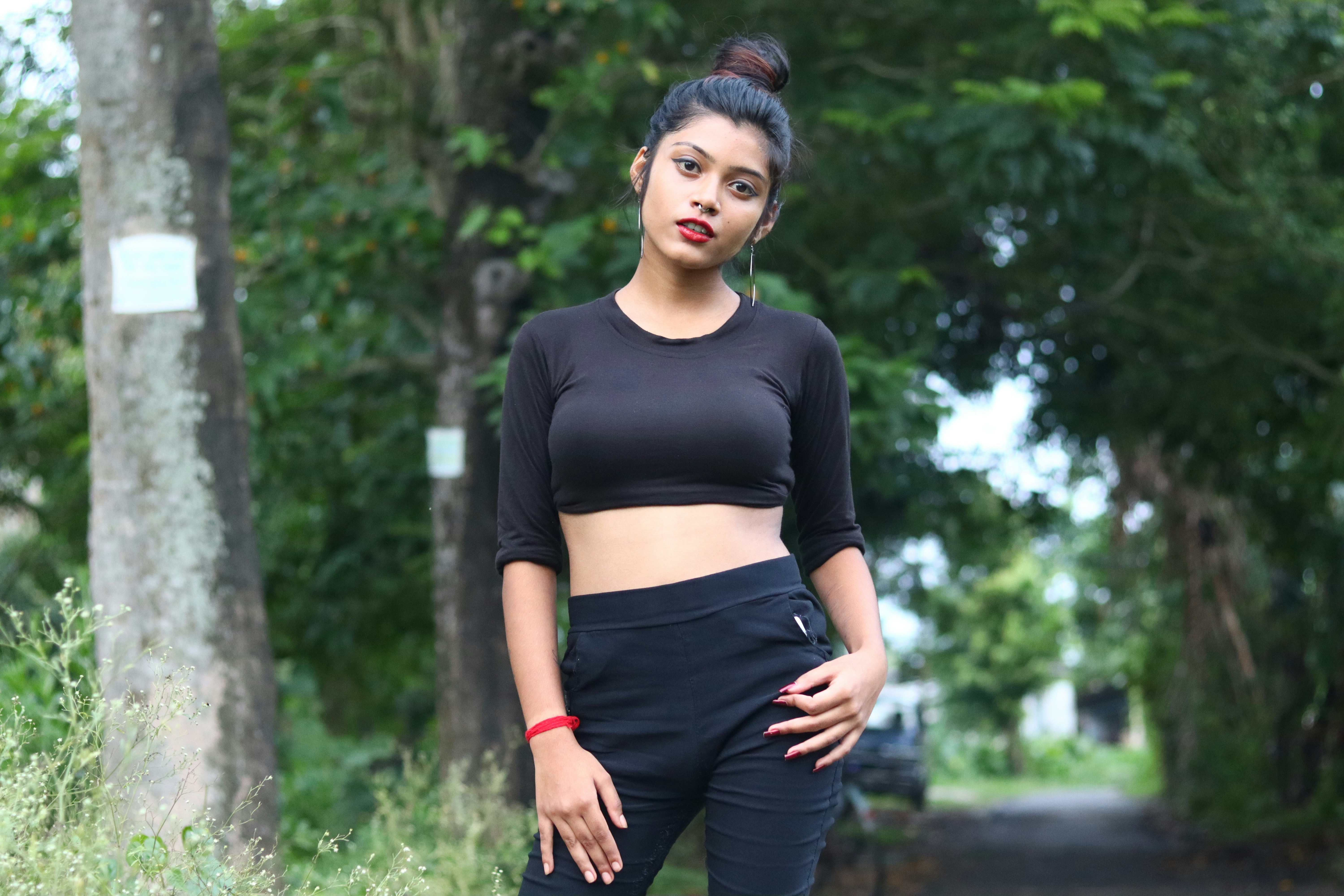 A young woman poses confidently in a black crop top and pants, framed by lush greenery and tree trunks along a pathway.