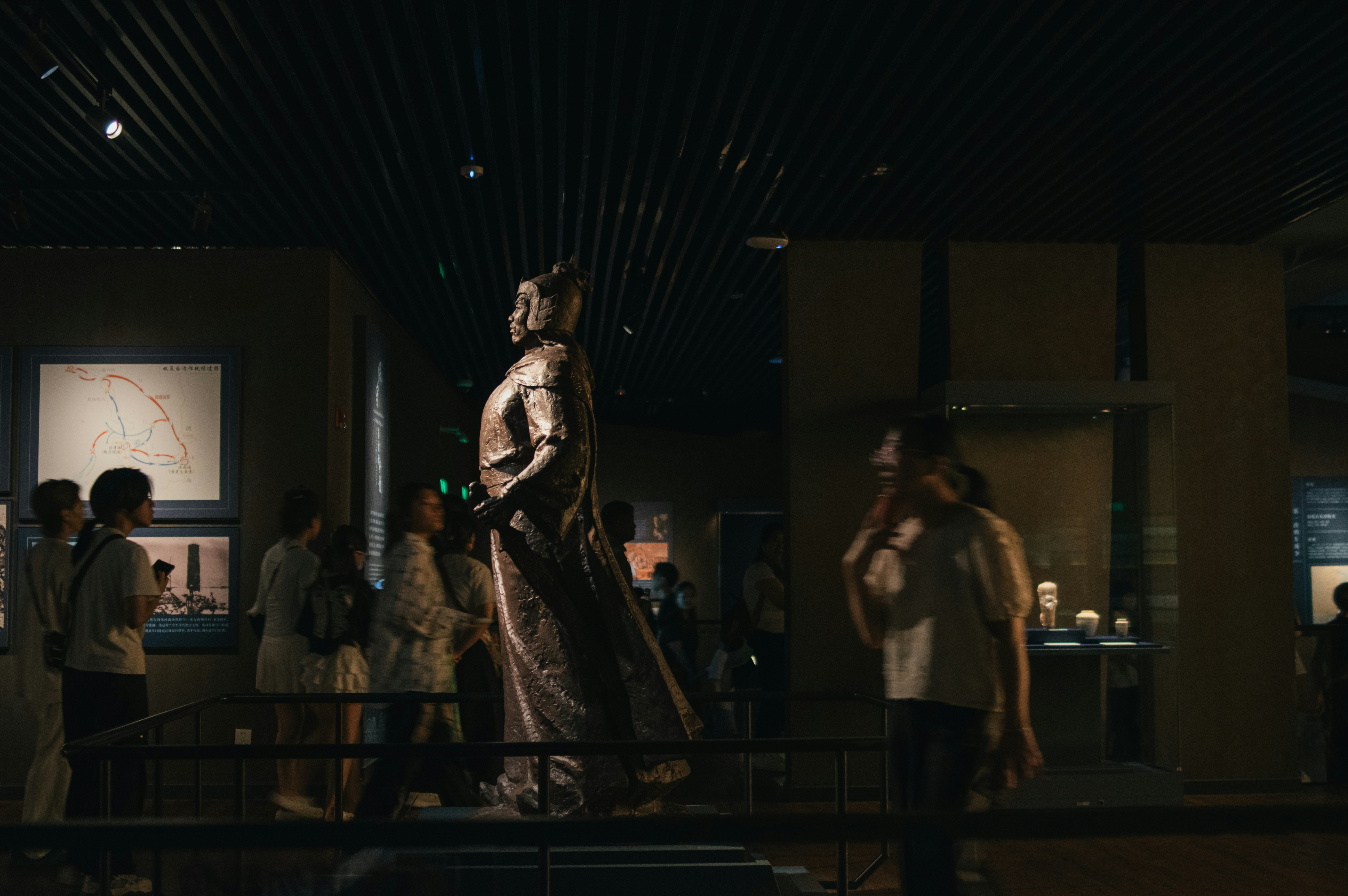 A beautifully crafted statue stands prominently in a museum, surrounded by visitors admiring the exhibits. The ambient lighting enhances the intricate details of the figure.