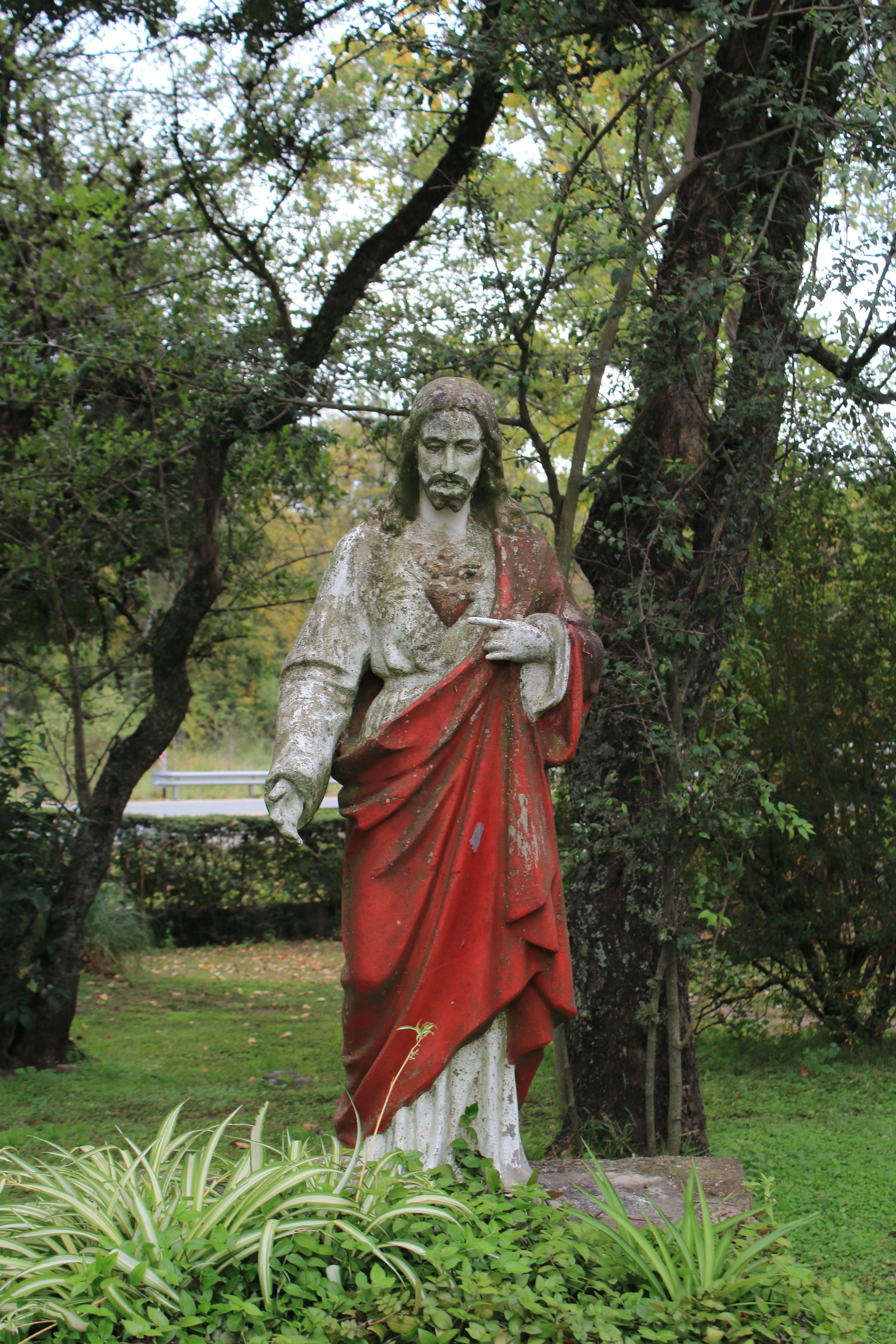 Weathered statue of a figure in red robes, standing among lush greenery, exuding a sense of tranquility and reflection.