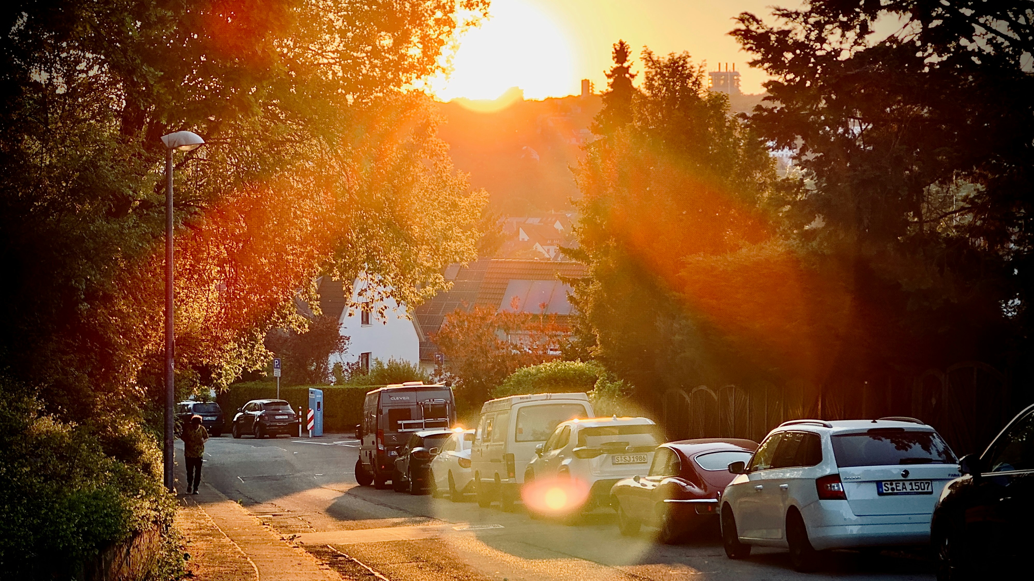 Sunset casting warm light over a tree-lined street, highlighting parked cars and a pedestrian. The scene captures the tranquility of evening light.
