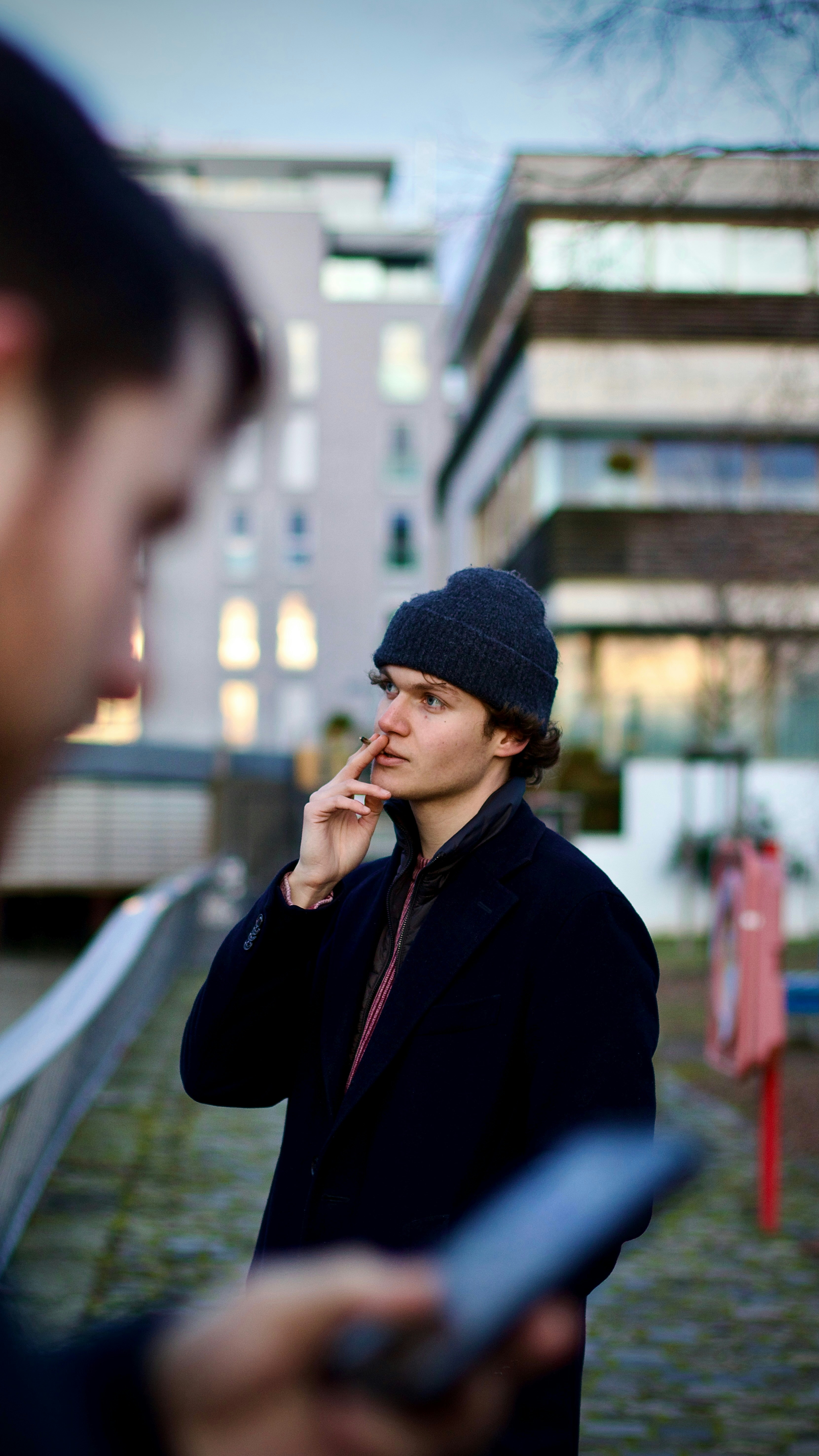 A young man in a beanie stands thoughtfully in an urban setting, with a blurred figure in the foreground holding a smartphone. The scene captures a moment of reflection against a modern backdrop.