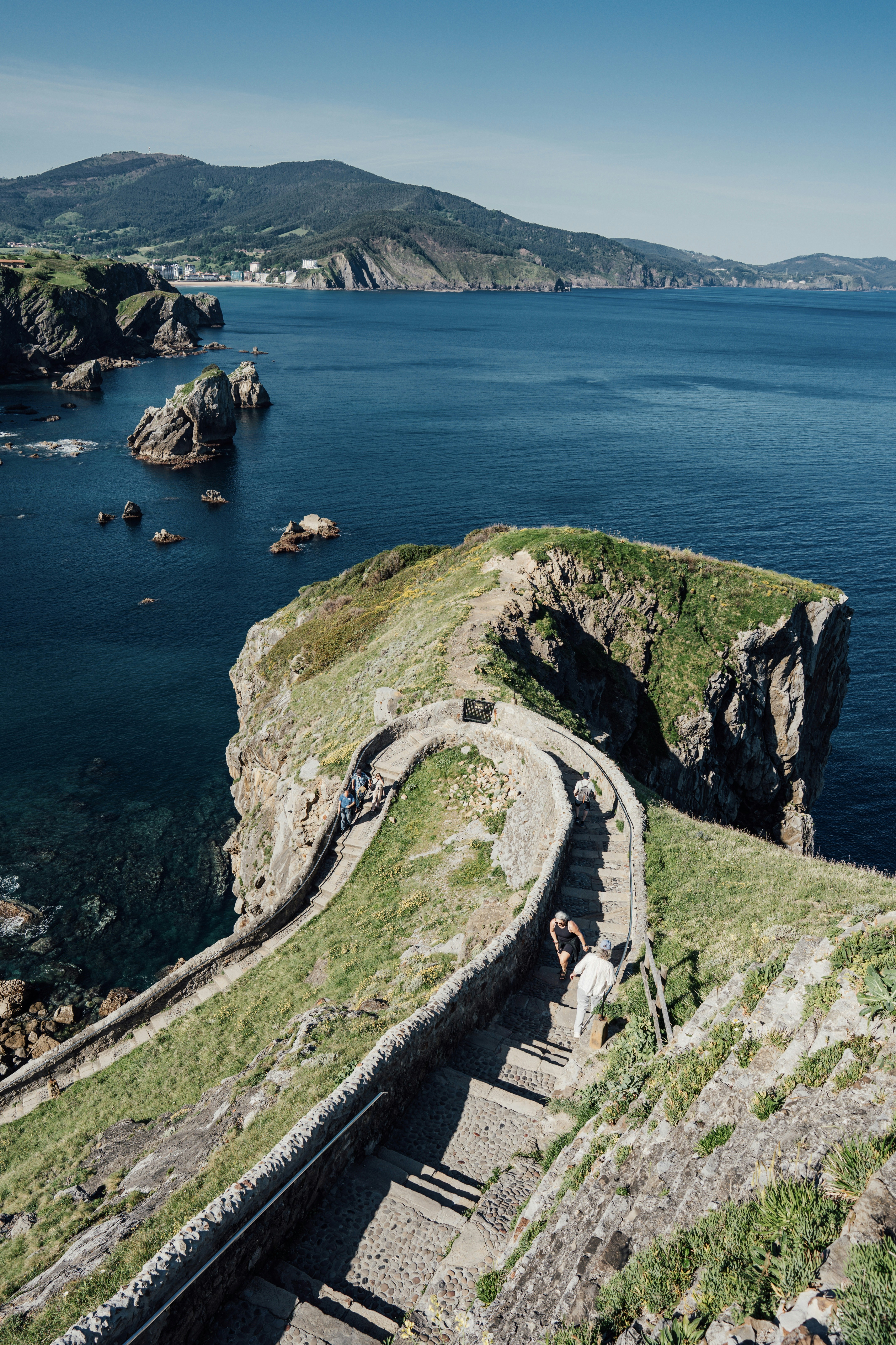 A steep, winding staircase leads down to a rocky coastline, surrounded by lush greenery and tranquil blue waters. The scene captures the harmony of nature and human exploration.