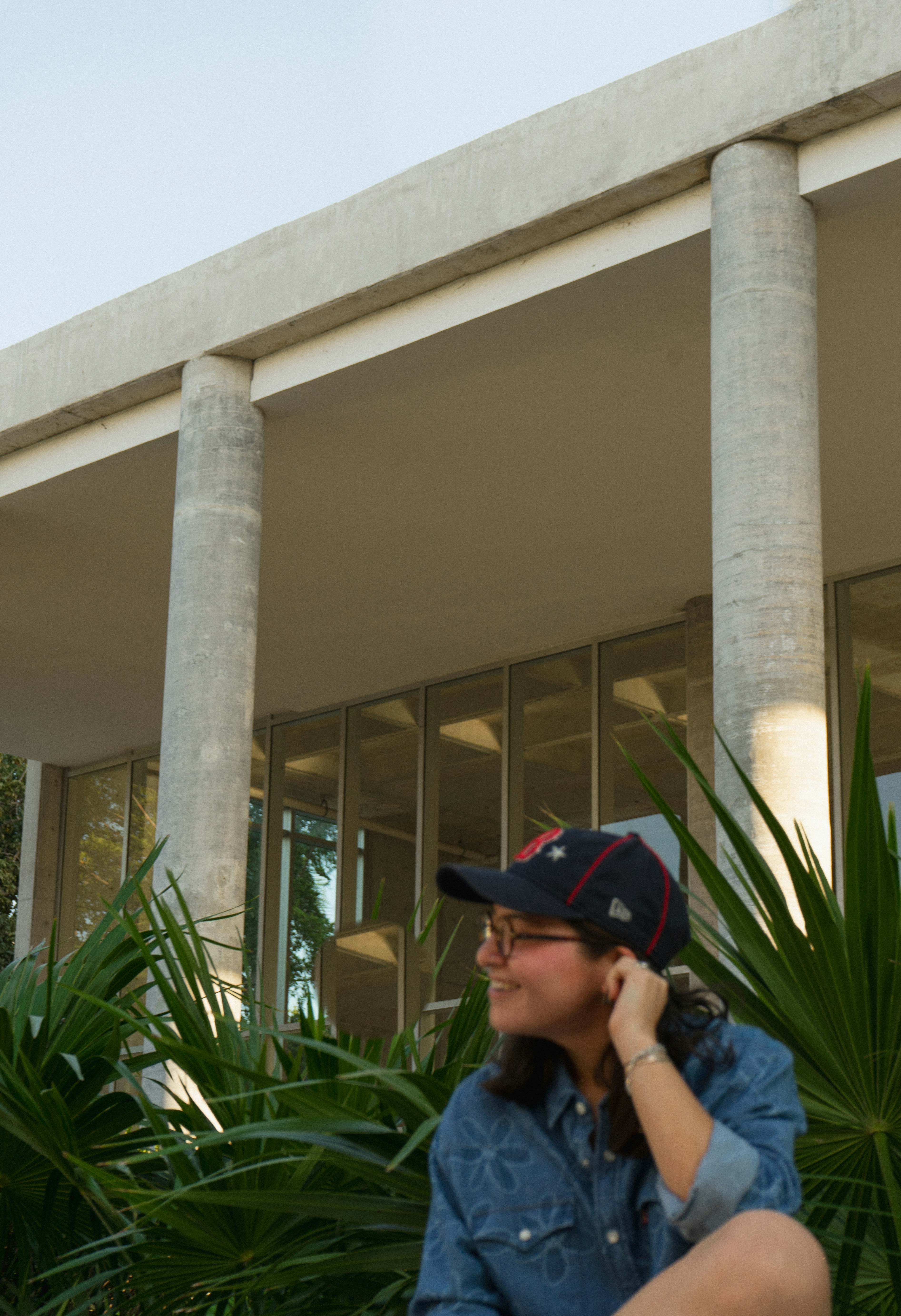 A young woman sitting among tropical plants, smiling softly, with a modern architectural backdrop. The scene embodies a blend of nature and contemporary design.