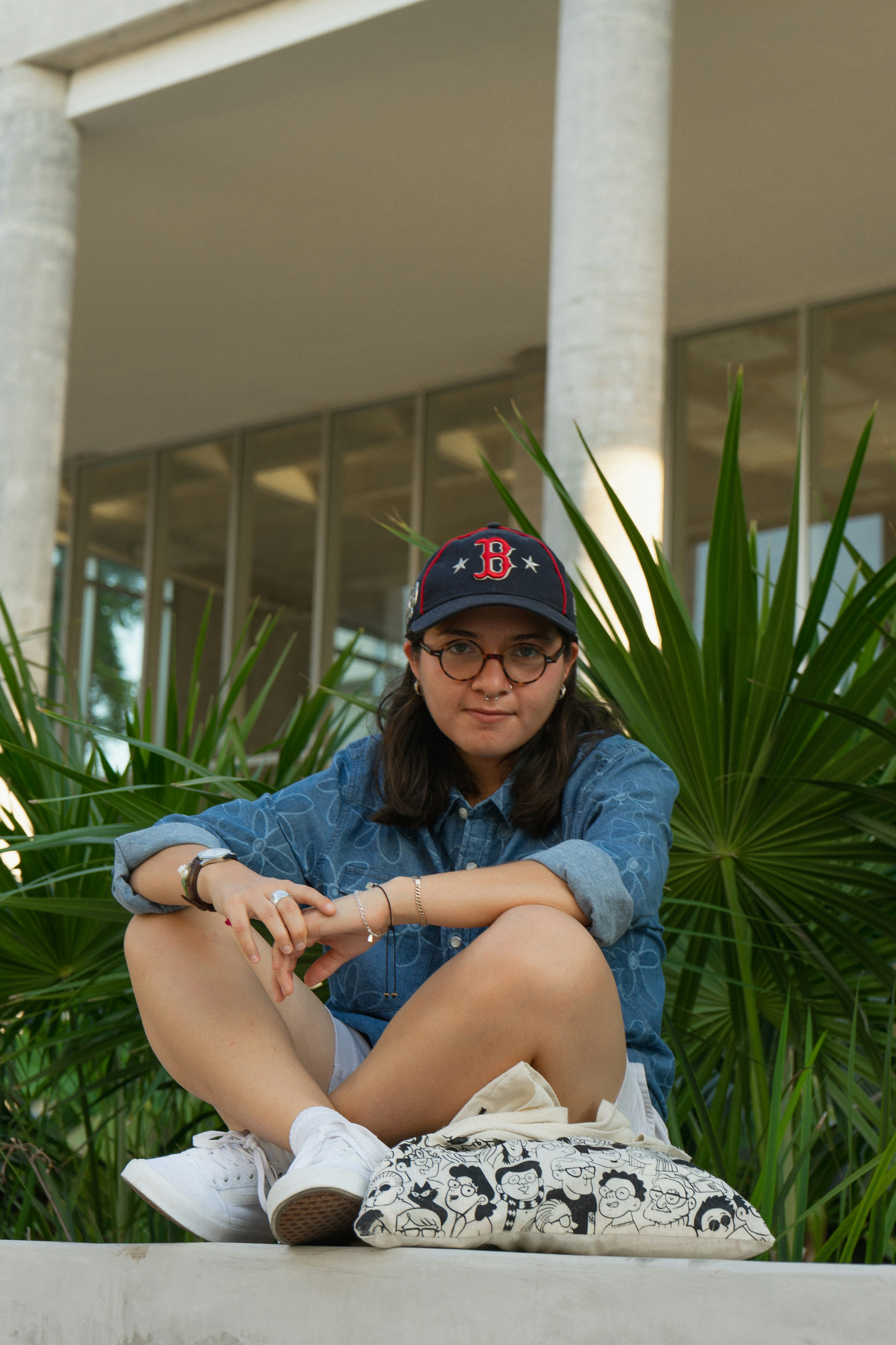 Young woman seated casually on a ledge surrounded by lush green plants, wearing a cap and denim shirt, exuding a relaxed vibe.