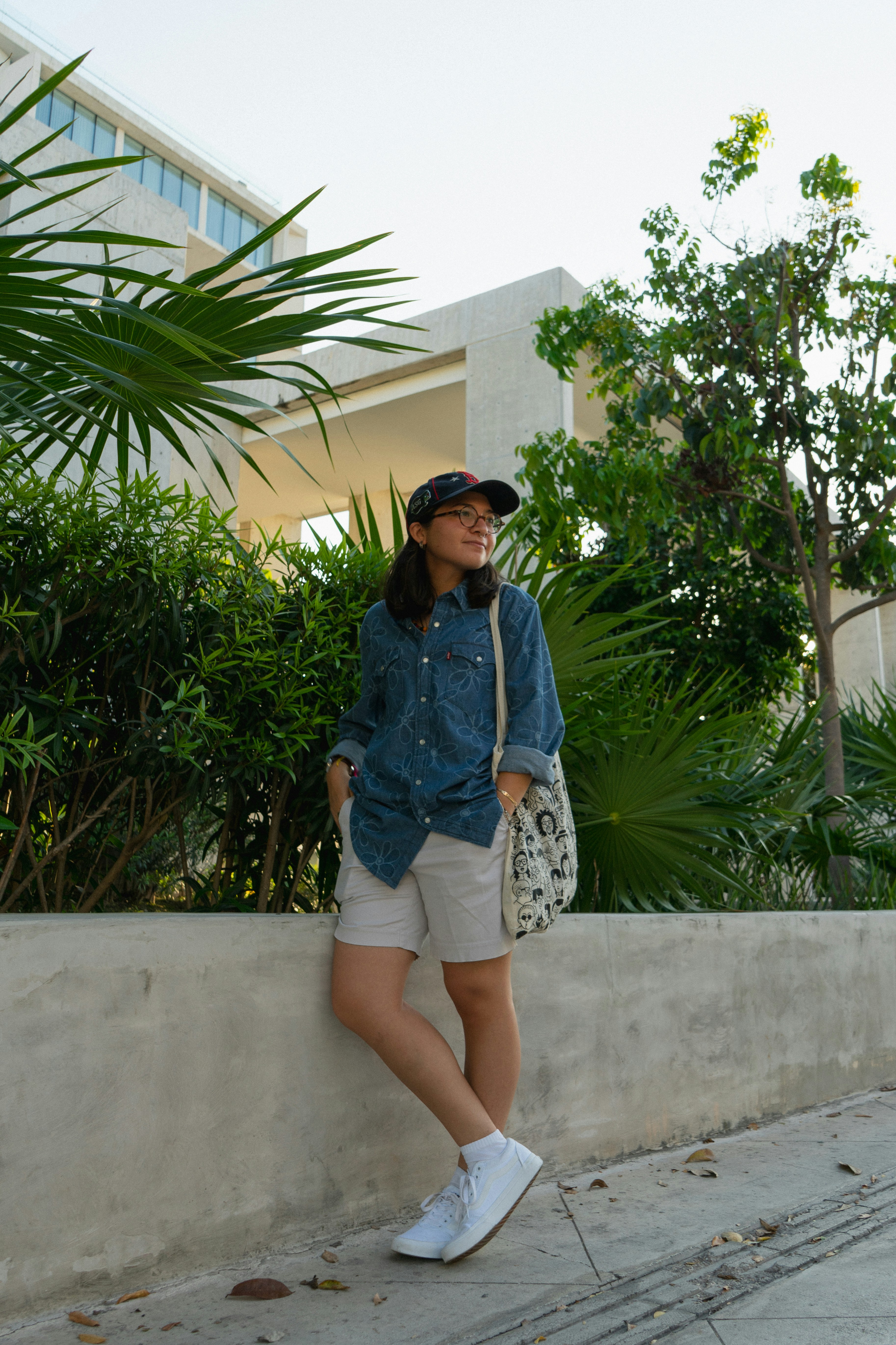 Young woman in casual attire stands against a low wall surrounded by lush plants, exuding a relaxed vibe in an urban setting.