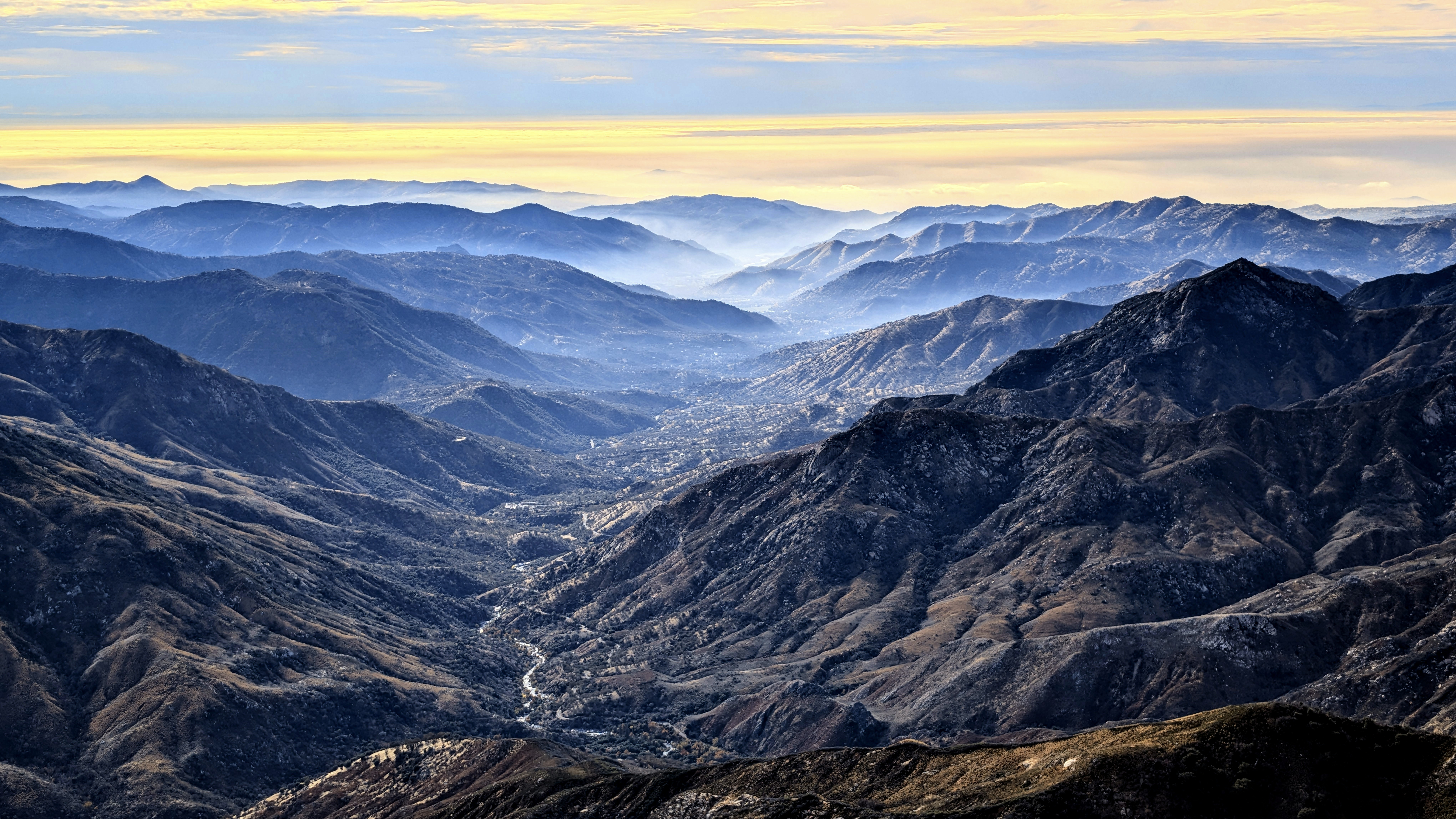 A sweeping view of layered mountain ranges shrouded in mist, revealing a winding valley below. The soft light casts a serene atmosphere.