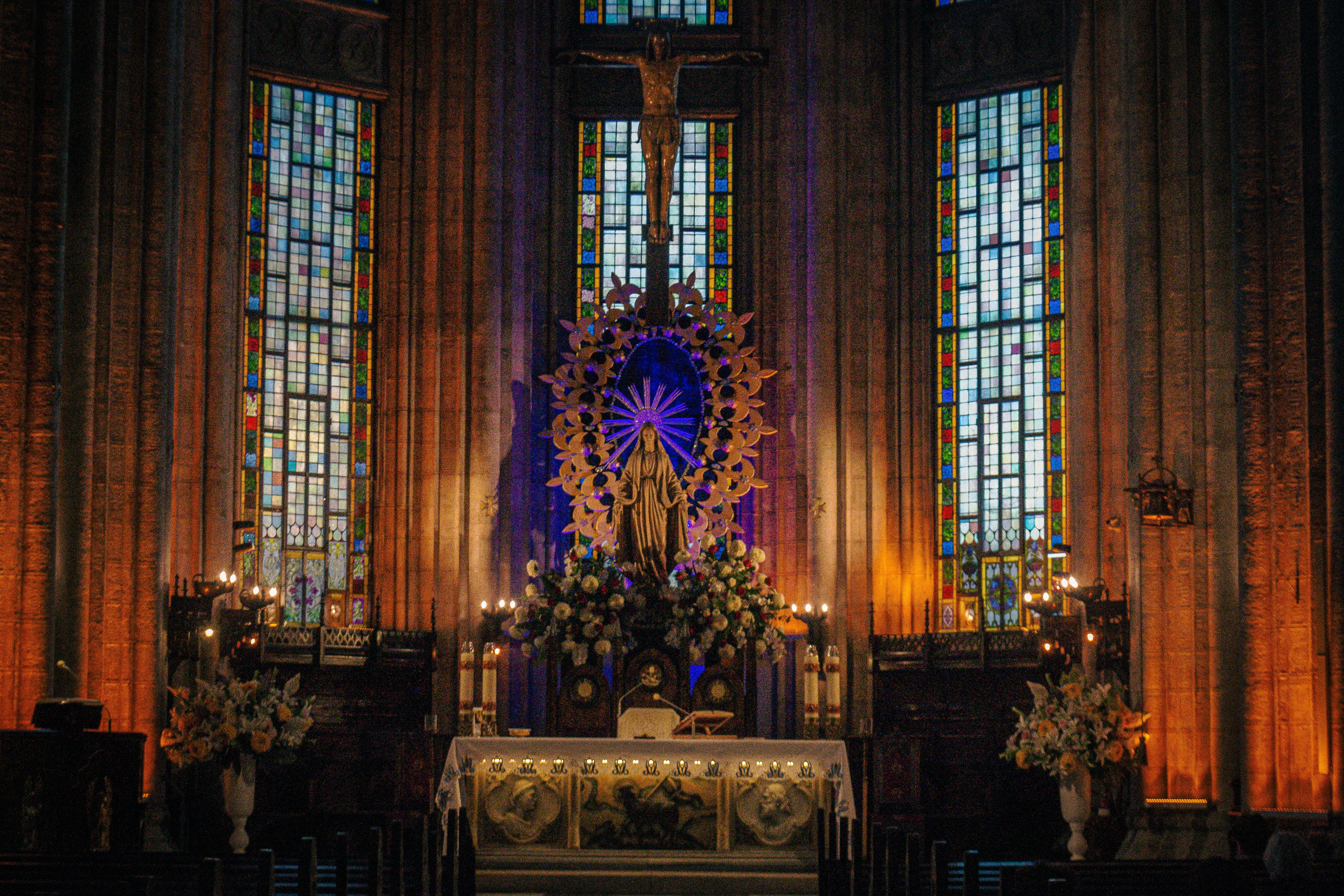 Altar adorned with flowers and candles in a church, featuring a statue surrounded by stained glass windows that cast colorful light.