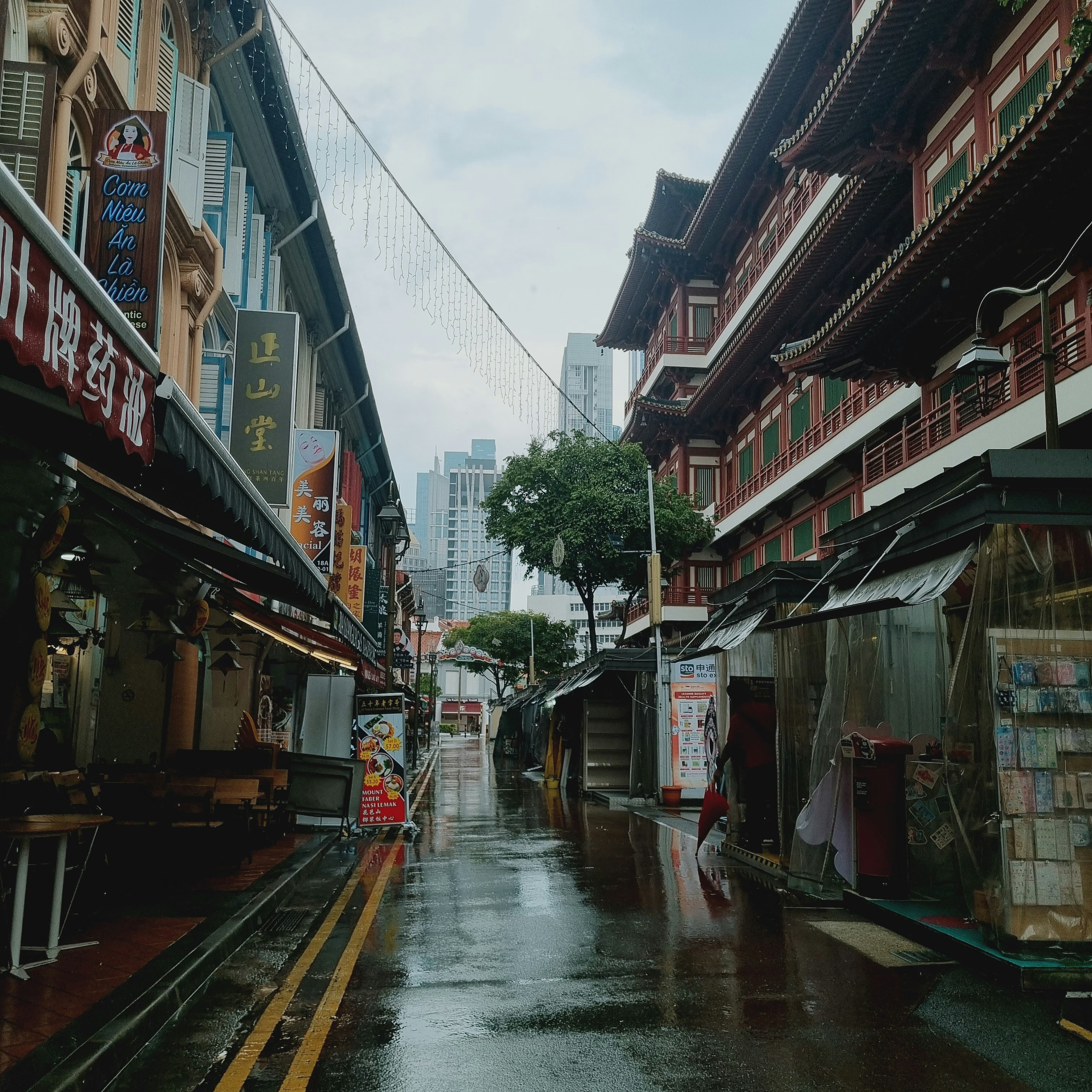 A vibrant street in Chinatown reflects the charm of traditional architecture, flanked by colorful stalls and modern skyscrapers. The wet pavement adds a serene atmosphere.