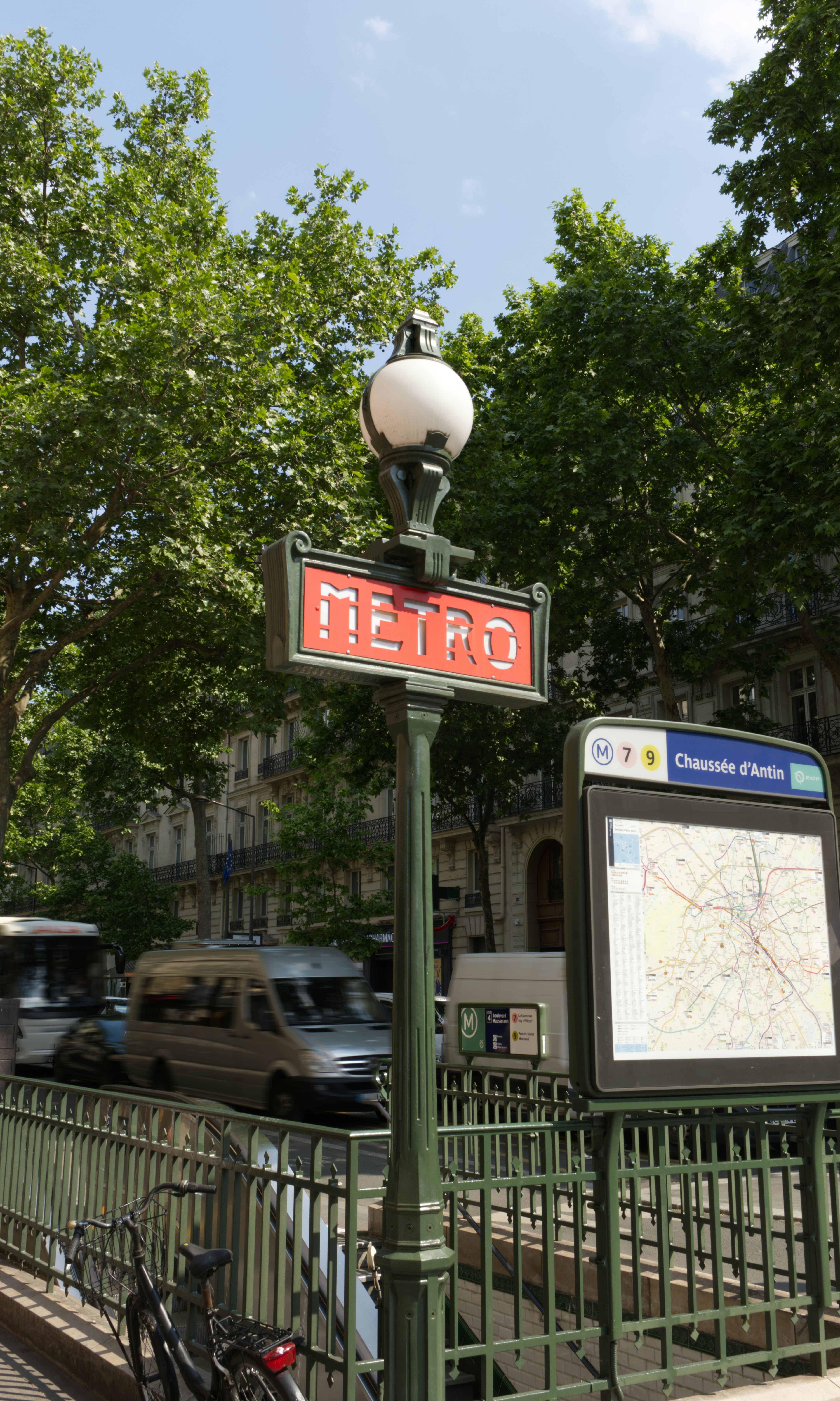 Historic METRO sign illuminated by sunlight, framed by lush green trees and urban architecture. Nearby transit map provides directional guidance.