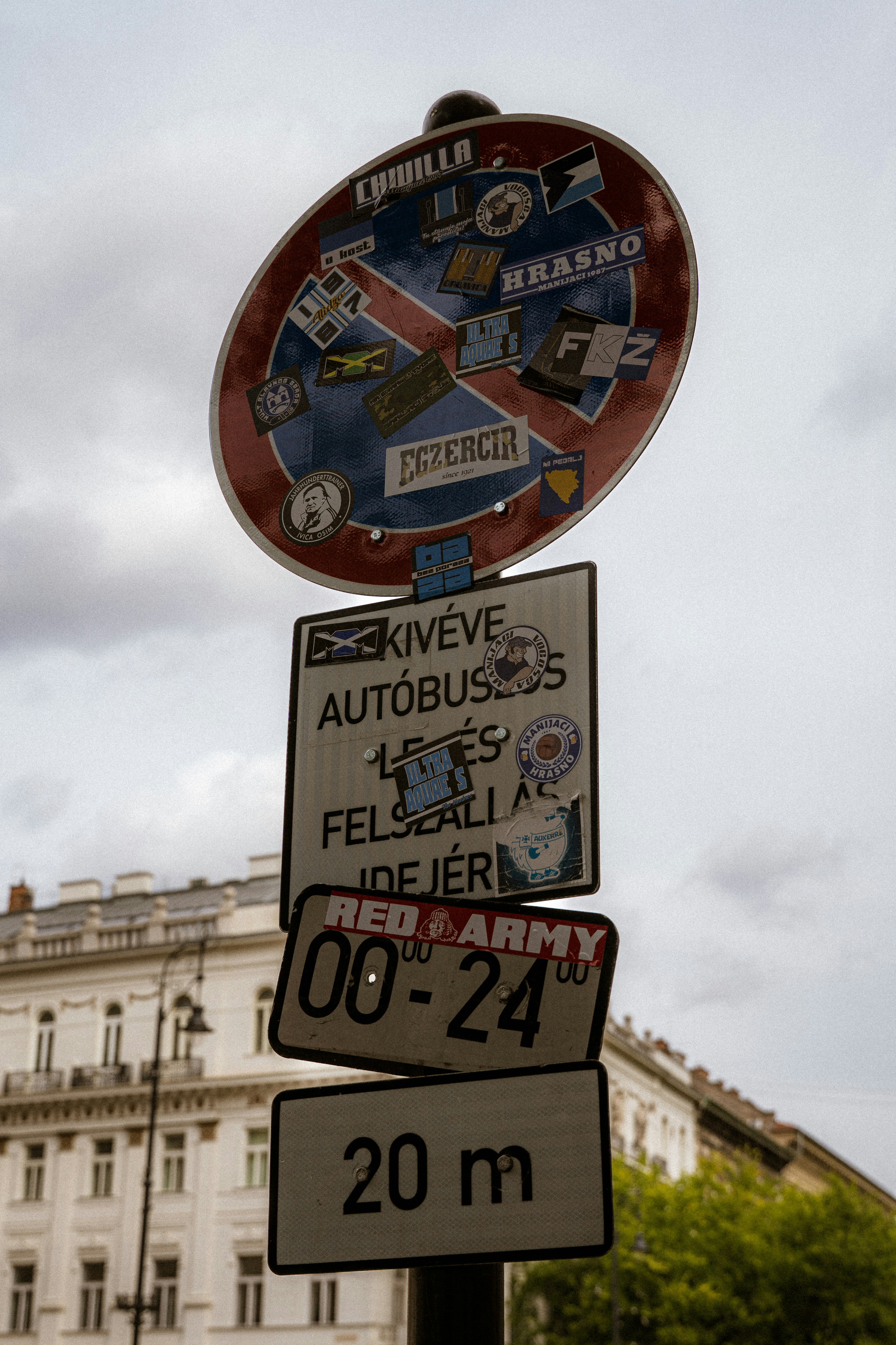 A bus stop sign adorned with various stickers and markings, showcasing a blend of urban culture and local identity. The backdrop features a cloudy sky and a historic building.