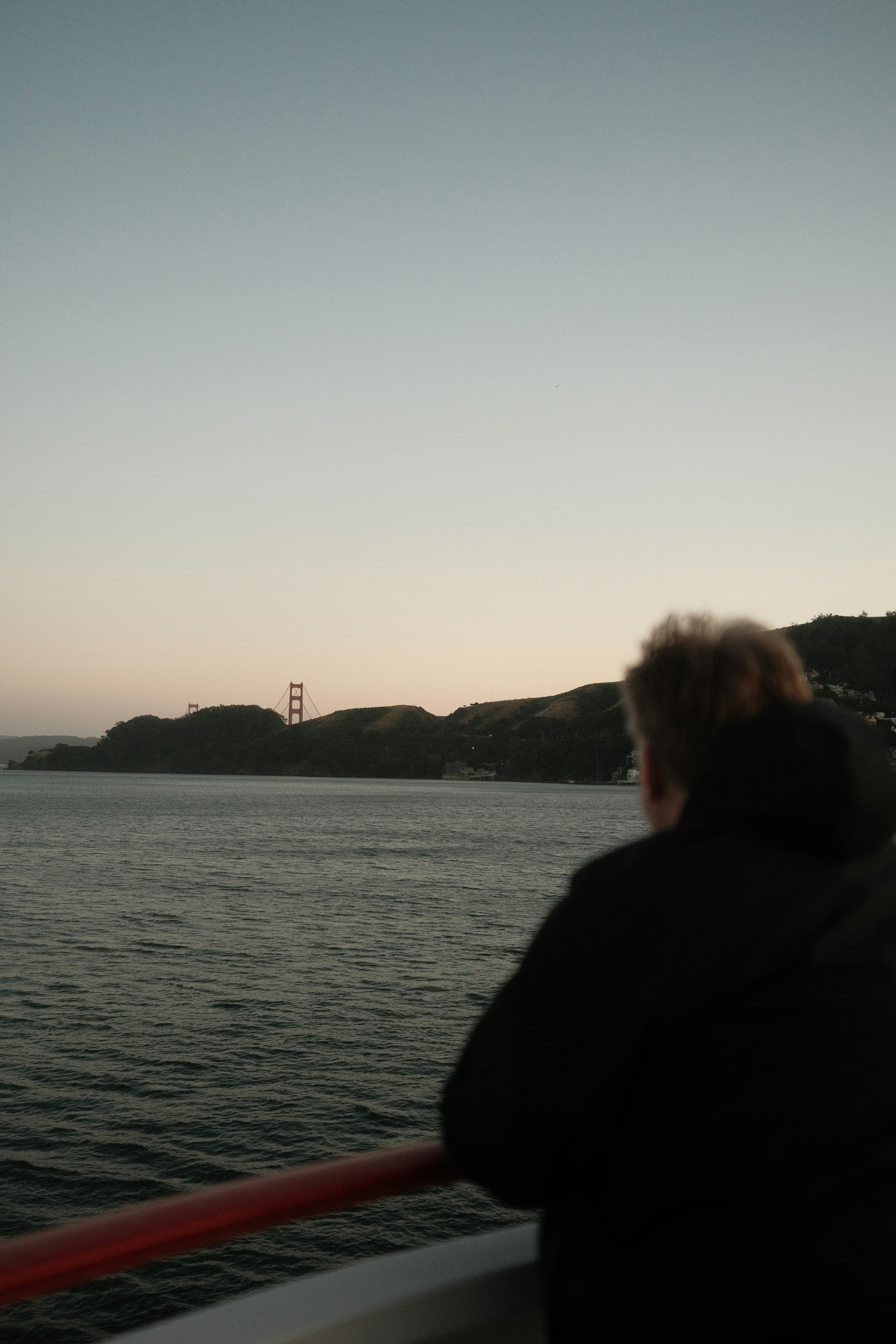 A person gazes thoughtfully at the Golden Gate Bridge from a boat, surrounded by tranquil waters at dusk.