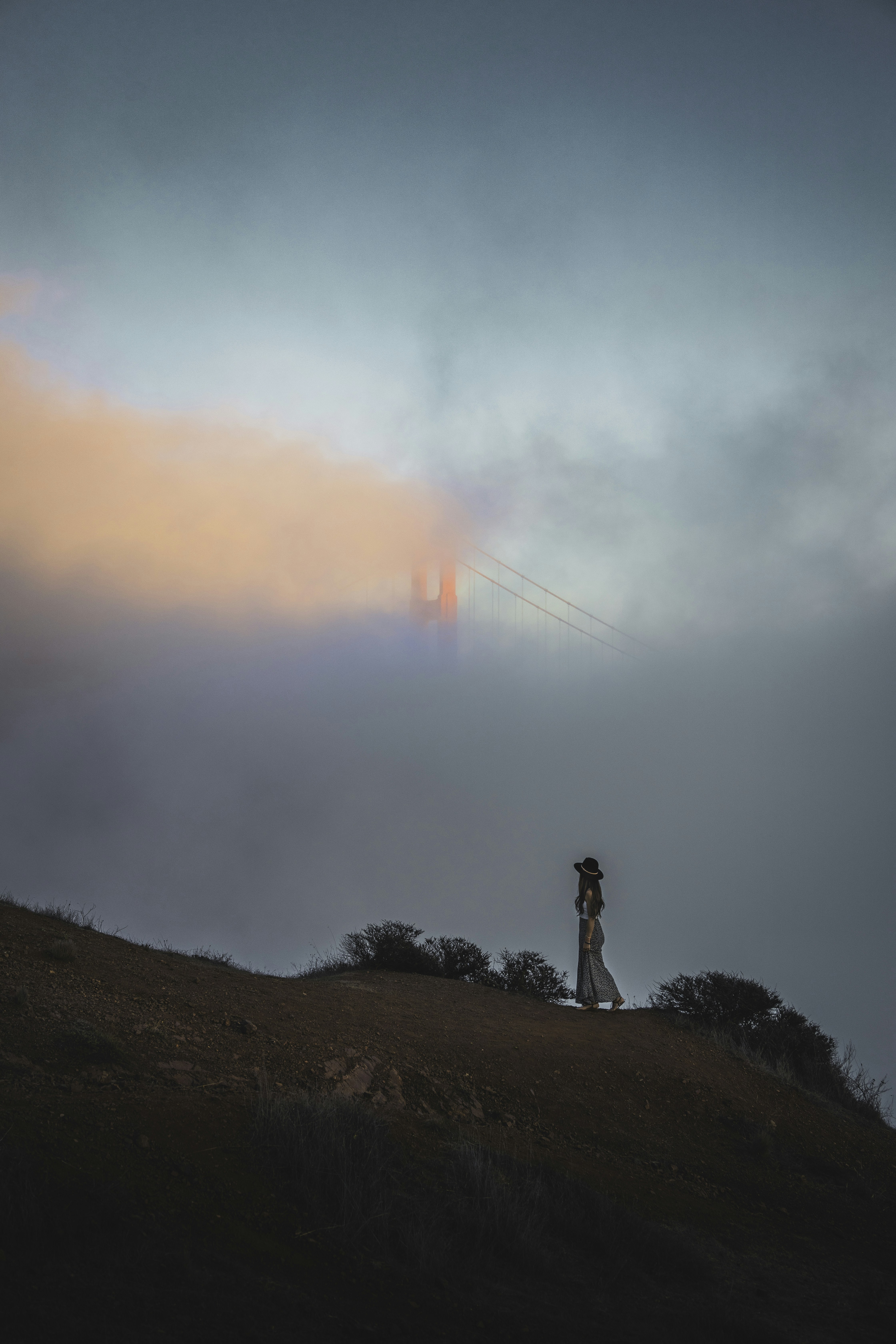 Silhouetted figure standing on a hillside, shrouded in fog with the Golden Gate Bridge faintly visible in the background. The scene evokes a sense of mystery and solitude.