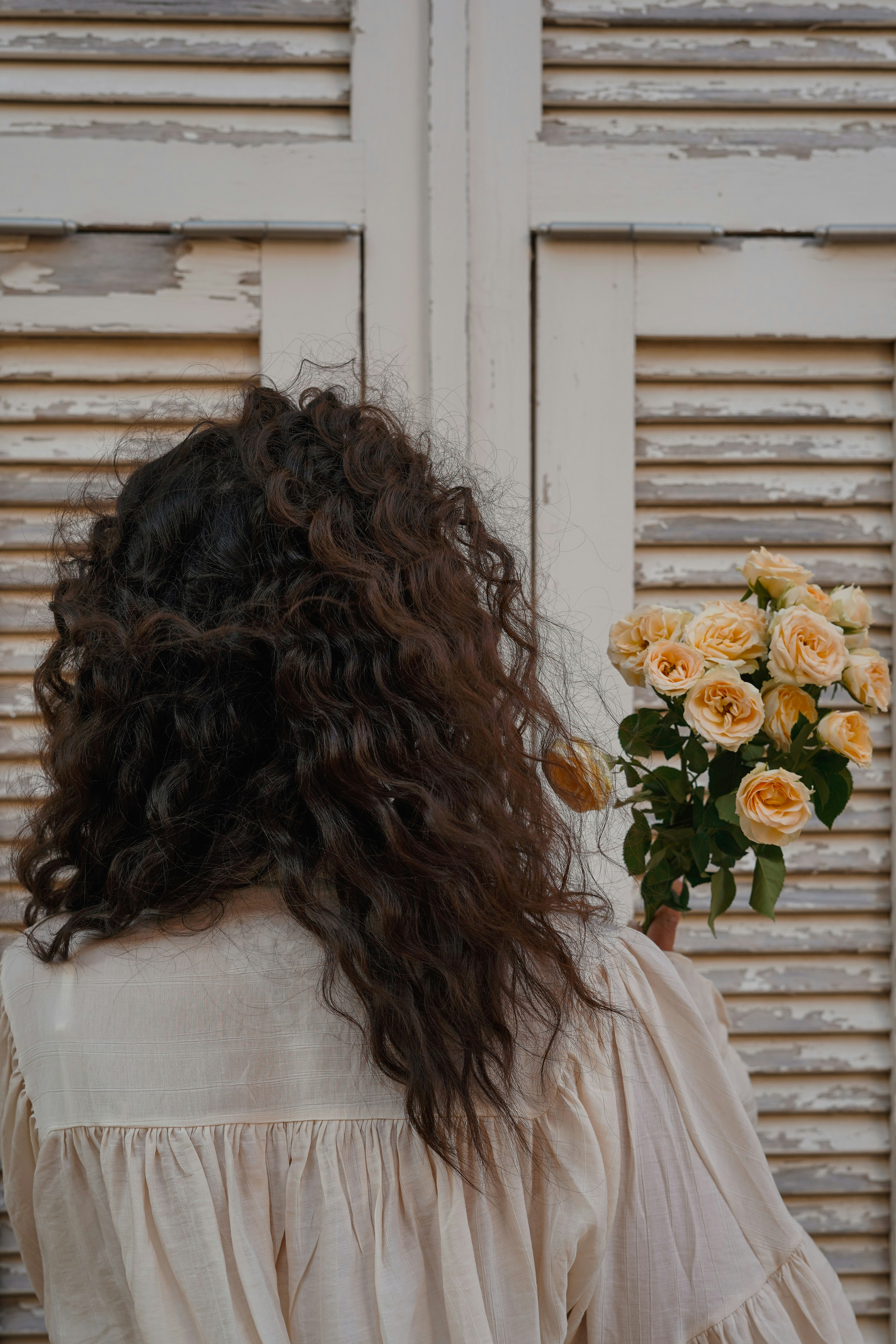 A woman with curly hair holds a bouquet of soft yellow roses against a textured, white shutter backdrop.
