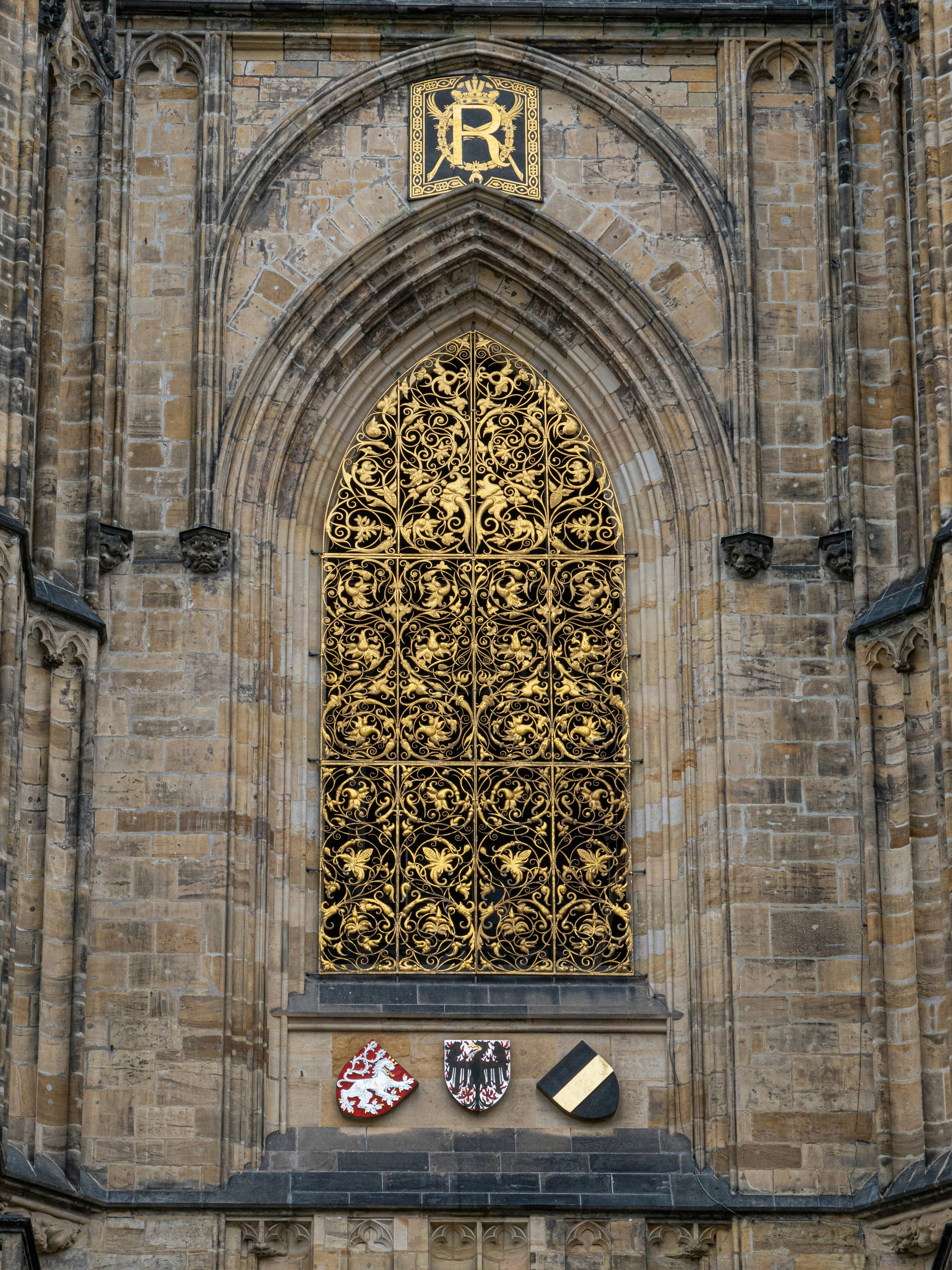 Intricately designed golden window featuring floral patterns, flanked by historical coats of arms on a stone facade.