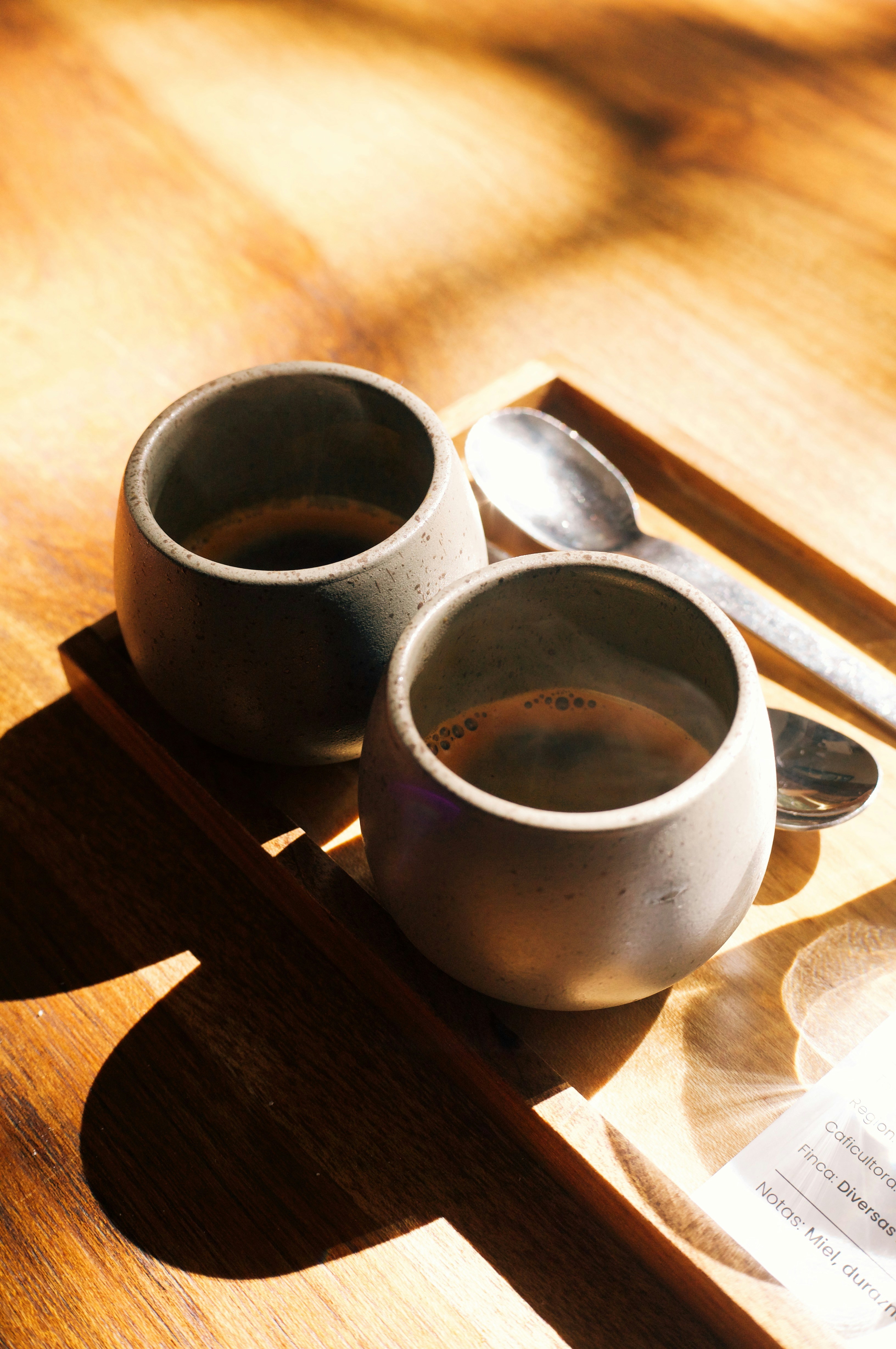 Two minimalist cups filled with coffee sit on a wooden tray, accompanied by a silver spoon and a small card detailing the brew. The warm sunlight casts soft shadows, enhancing the serene atmosphere.