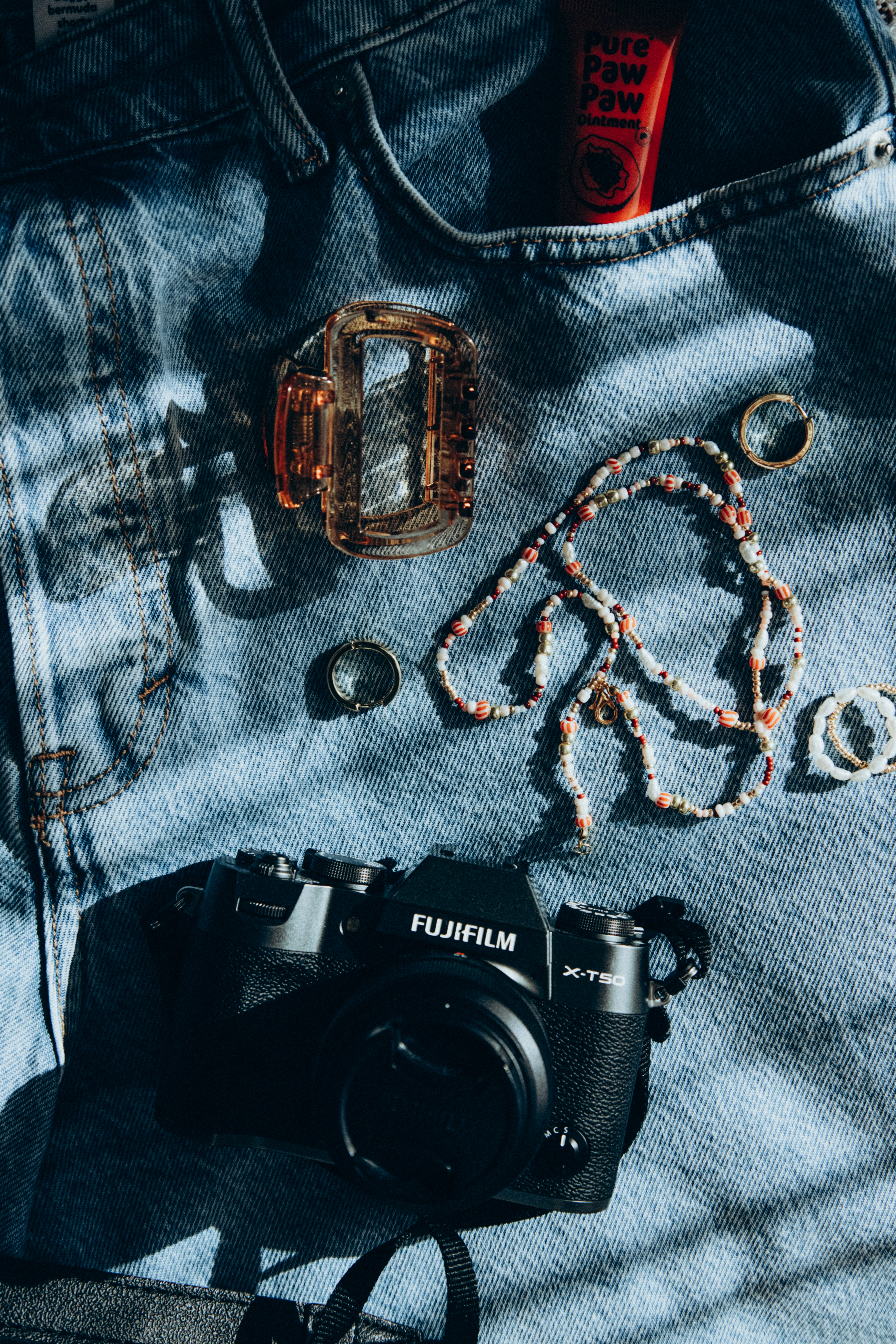 A Fujifilm X-T30 camera rests on a pair of denim jeans, surrounded by accessories like a hair clip and beaded necklace, highlighting a laid-back, artistic lifestyle.