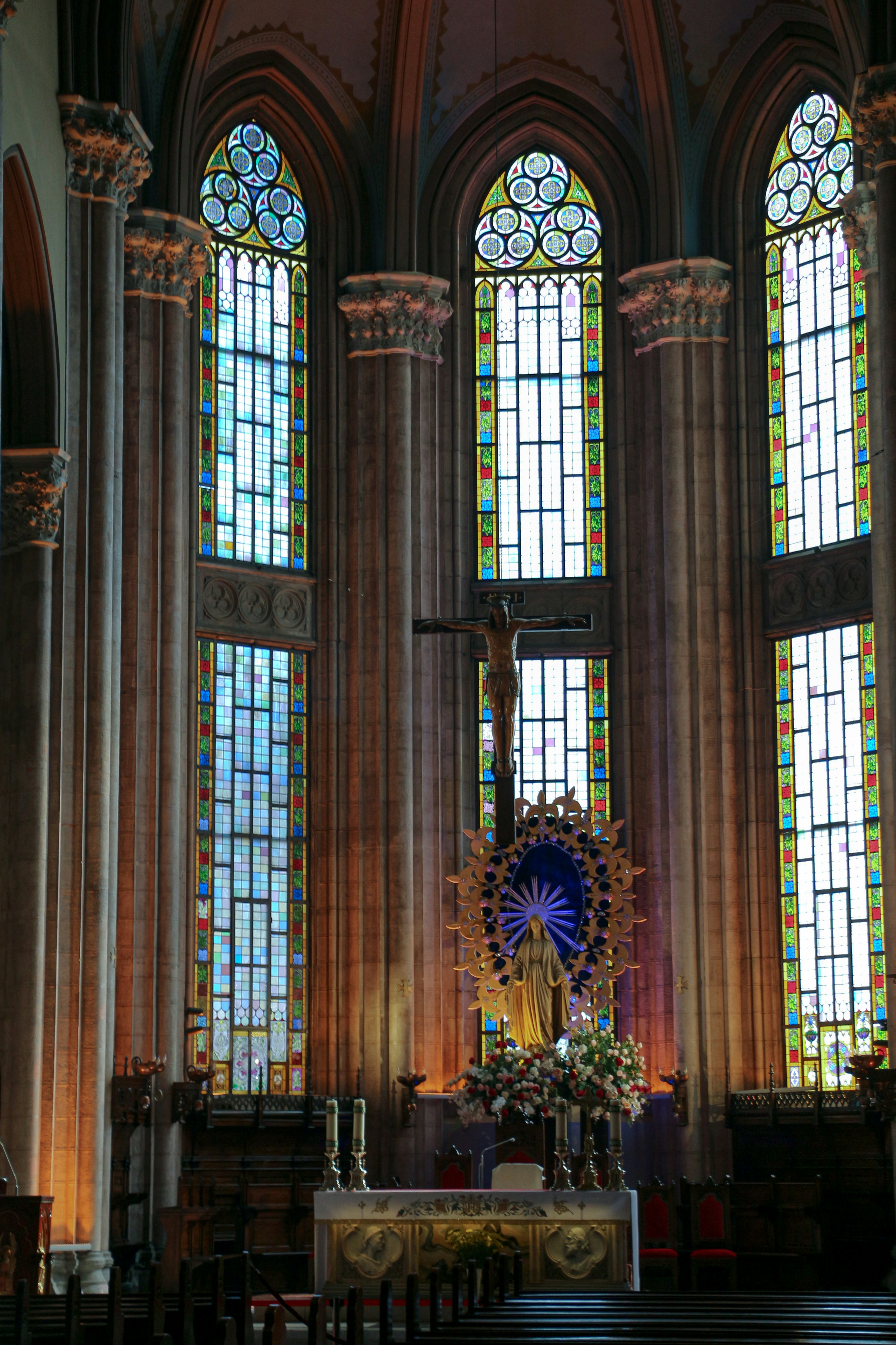 Intricate stained glass windows illuminate the altar, showcasing a statue of the Virgin Mary surrounded by floral arrangements. The warm tones create a serene atmosphere.